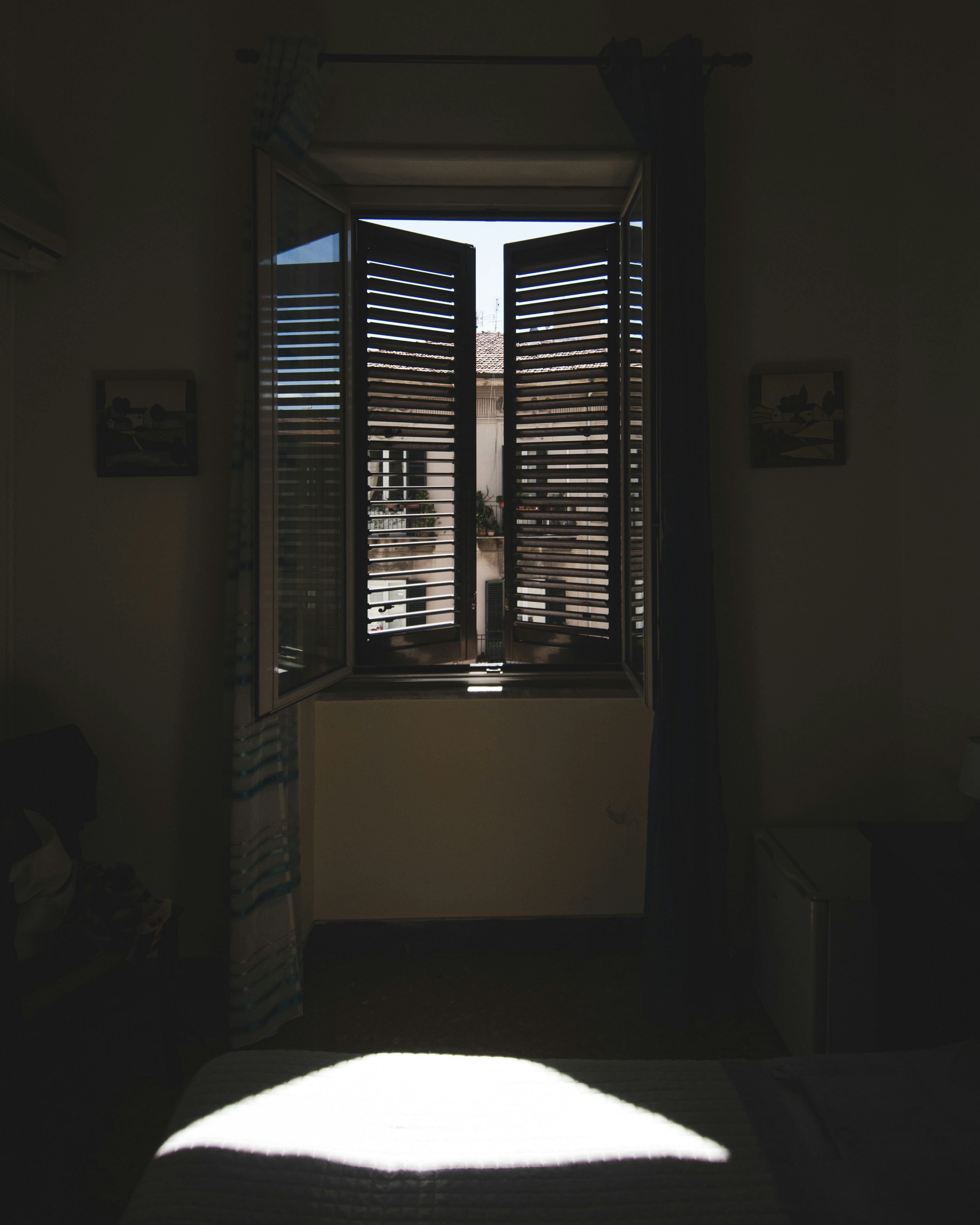 Open window with wooden shutters casting shadows in a dimly lit room, revealing a glimpse of the outside world. 