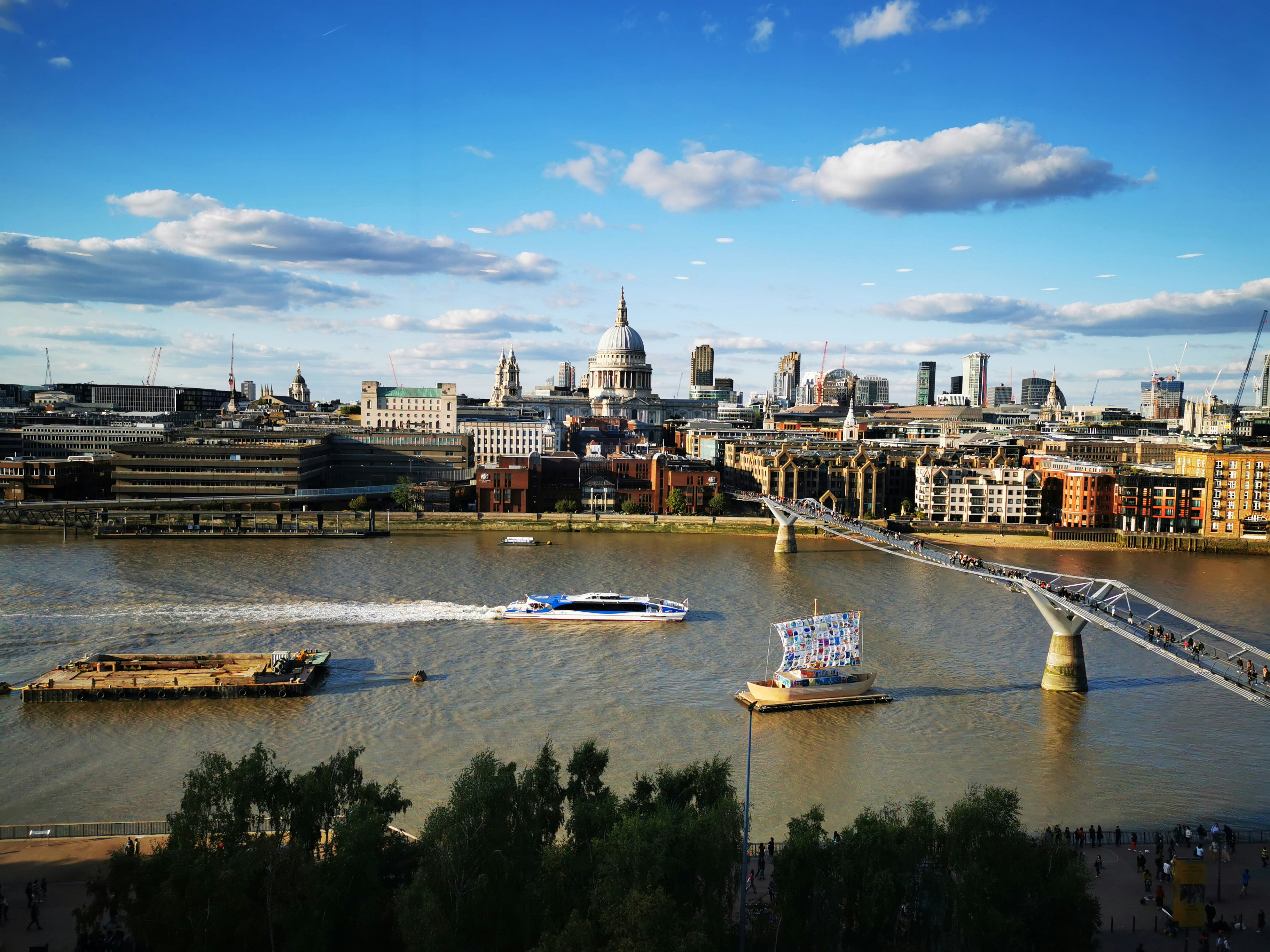A panoramic view of the Thames River with St. Paul's Cathedral in the background, showcasing boats navigating the water and pedestrians on the Millennium Bridge.