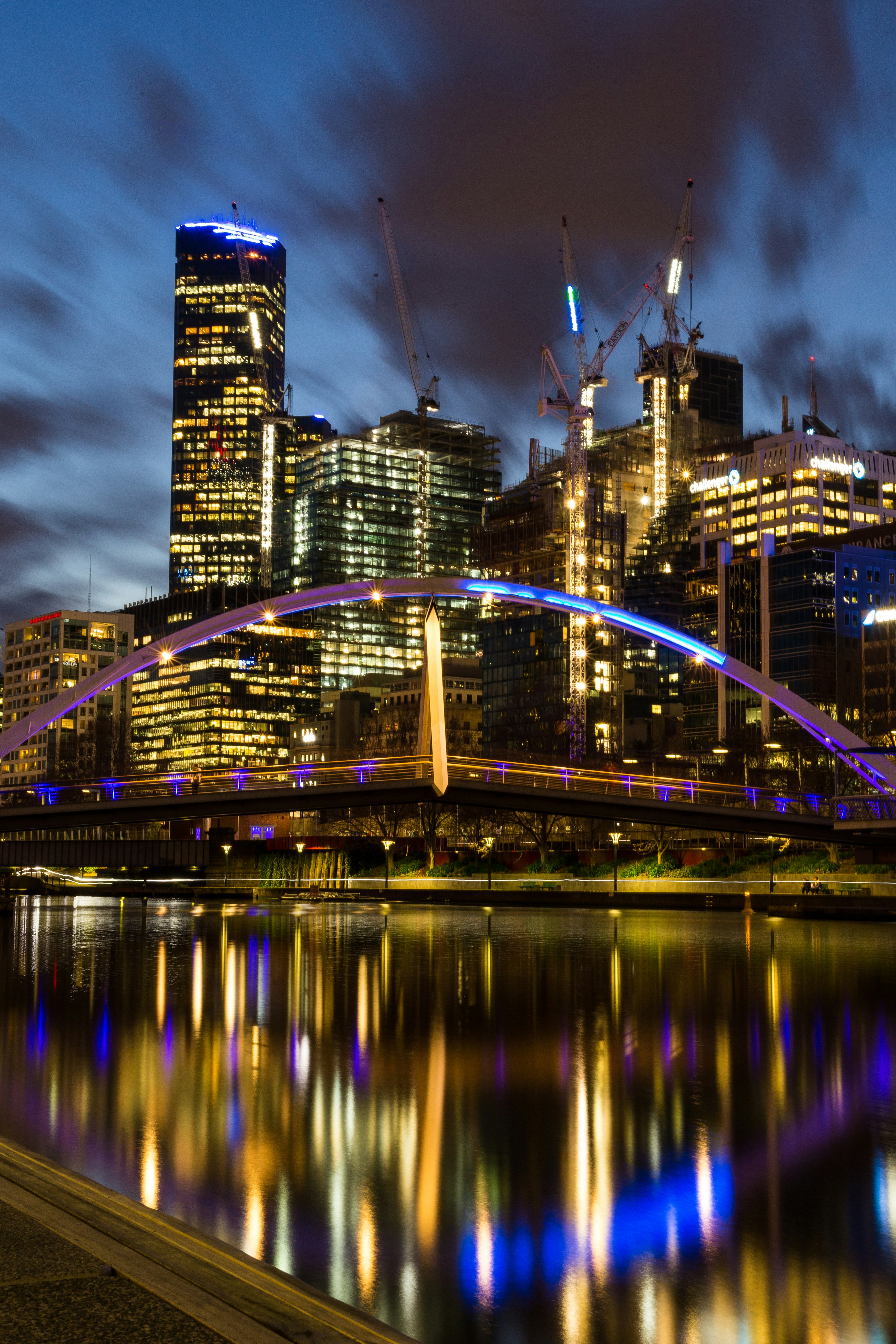 Illuminated city skyline reflecting on the river at dusk, featuring a striking bridge and ongoing construction. The scene captures the vibrant energy of urban development.