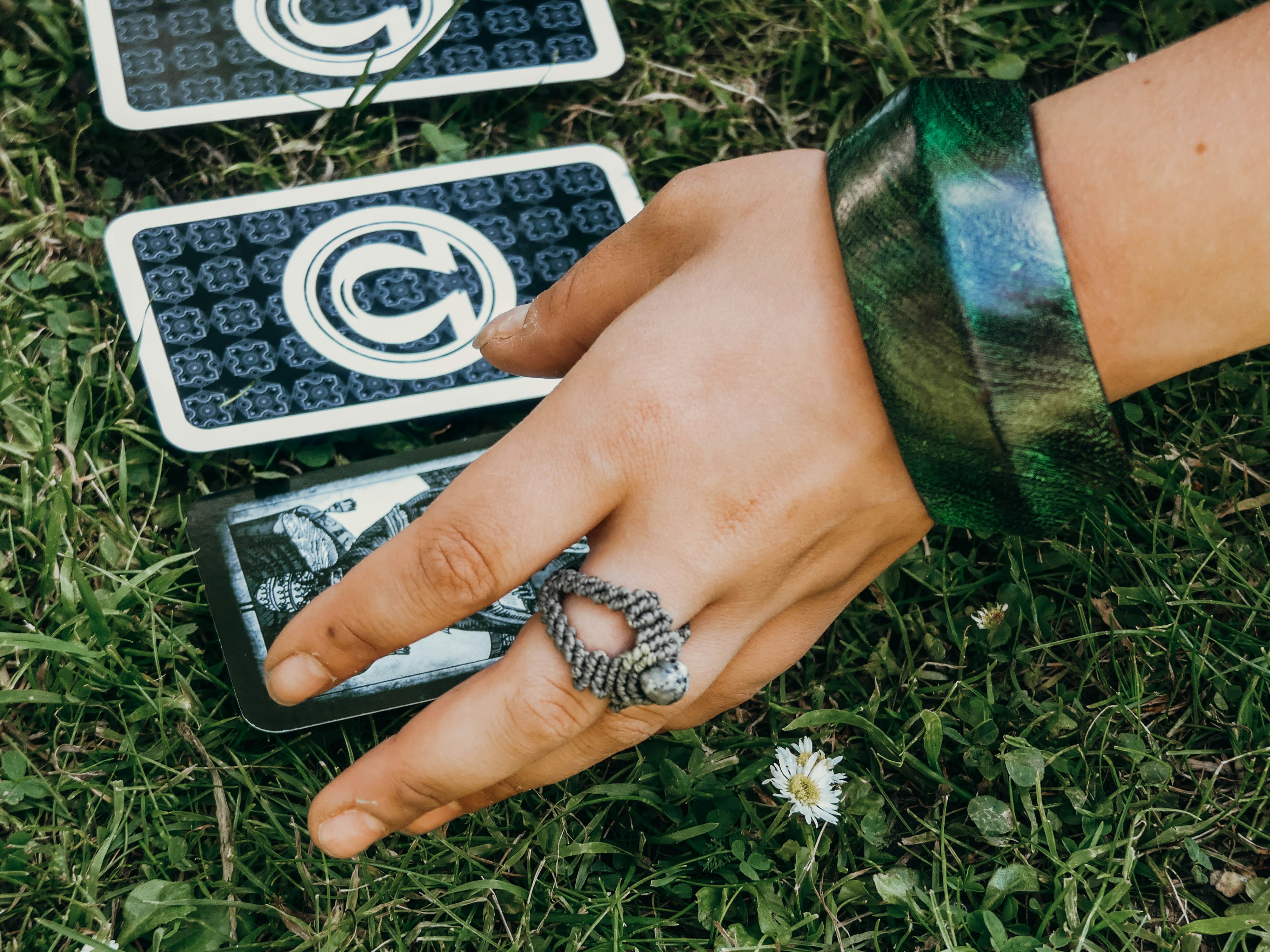 A hand adorned with a silver ring and a green bracelet interacts with playing cards on grass, suggesting a moment of contemplation or decision.
