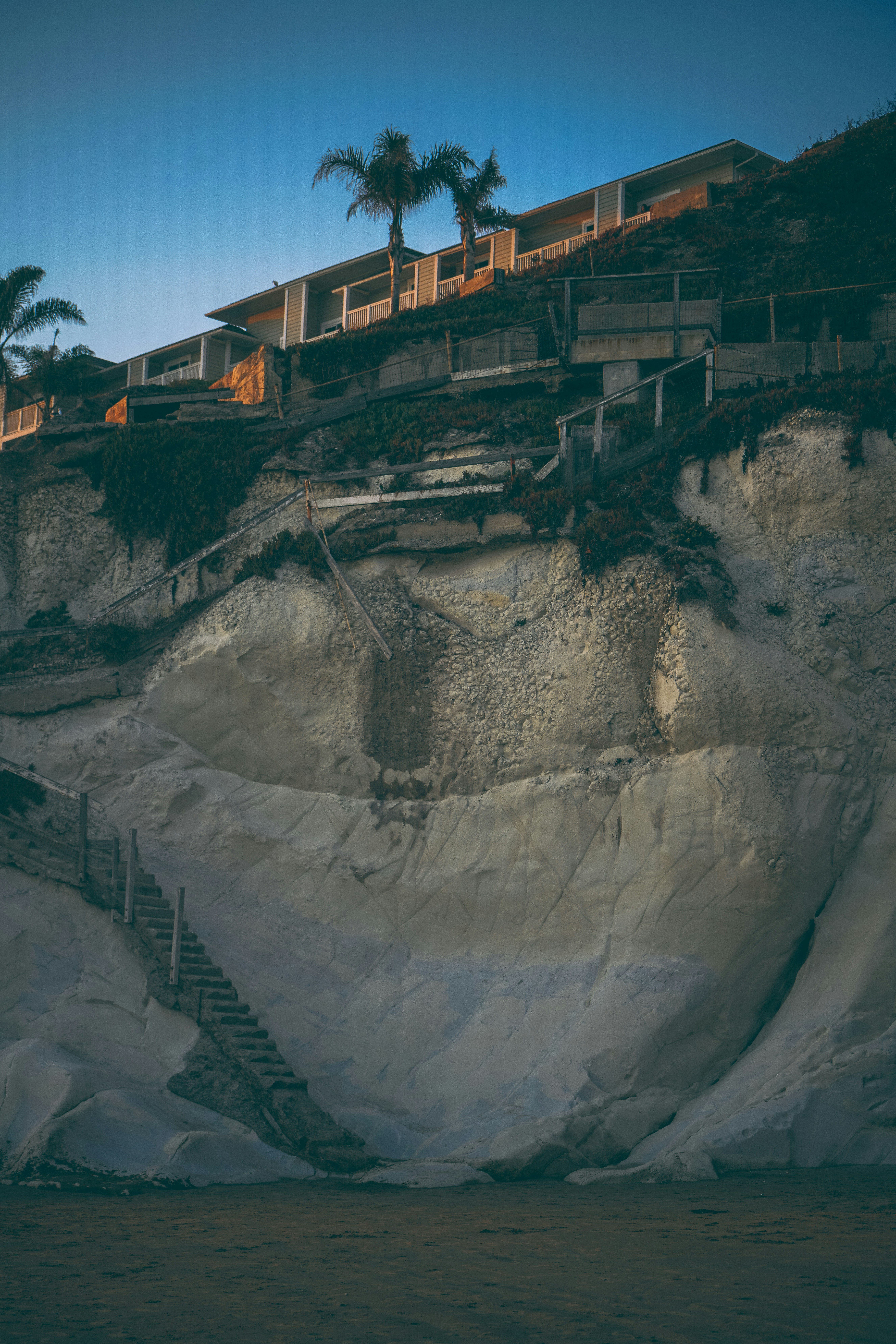 A coastal cliff showcasing a modern home perched above, surrounded by palm trees and rugged terrain. The intricate layers of the cliff reveal natural textures and colors.