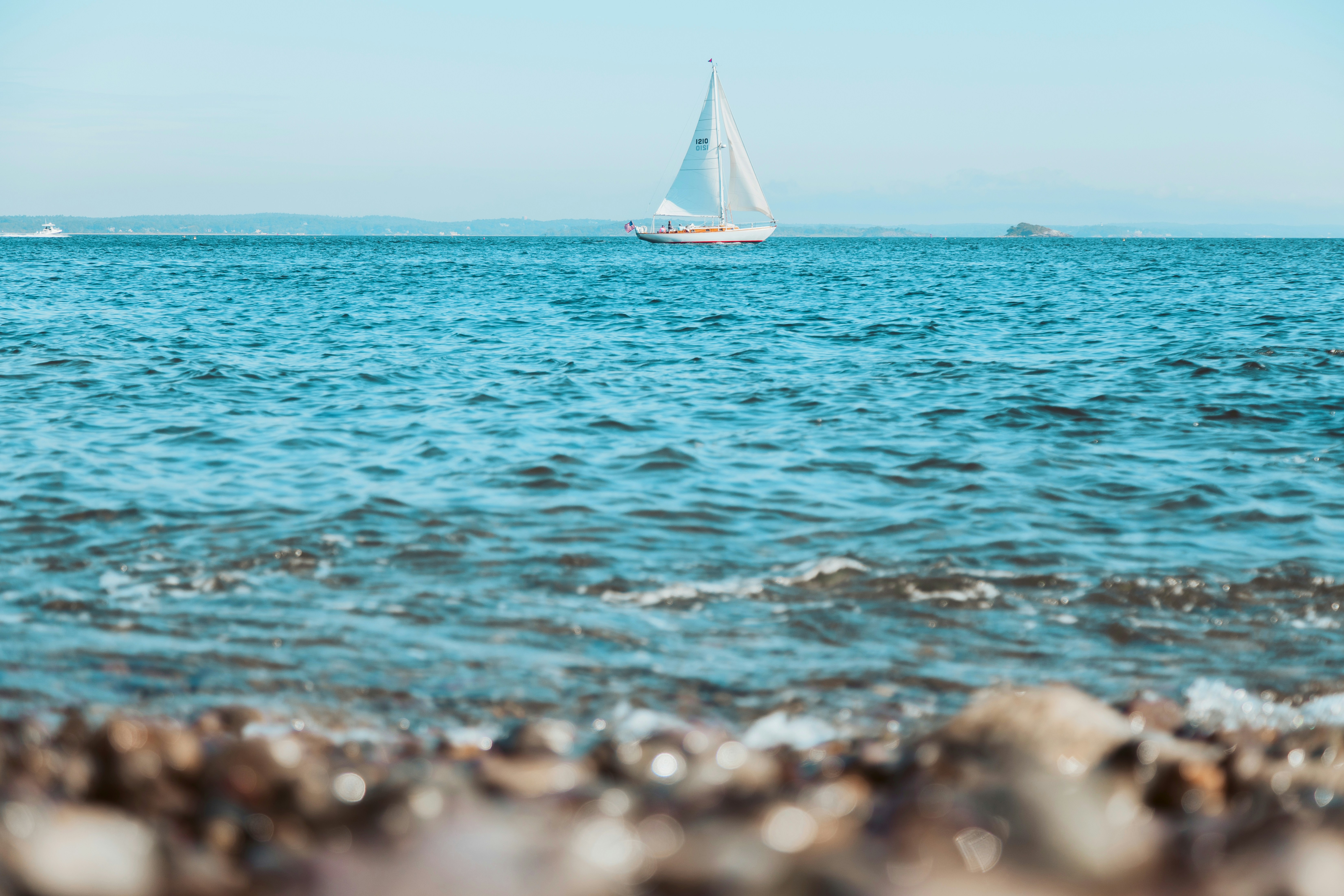 A sailboat glides across a tranquil sea, framed by a rocky shoreline and a clear sky. The scene evokes a sense of calm and adventure.