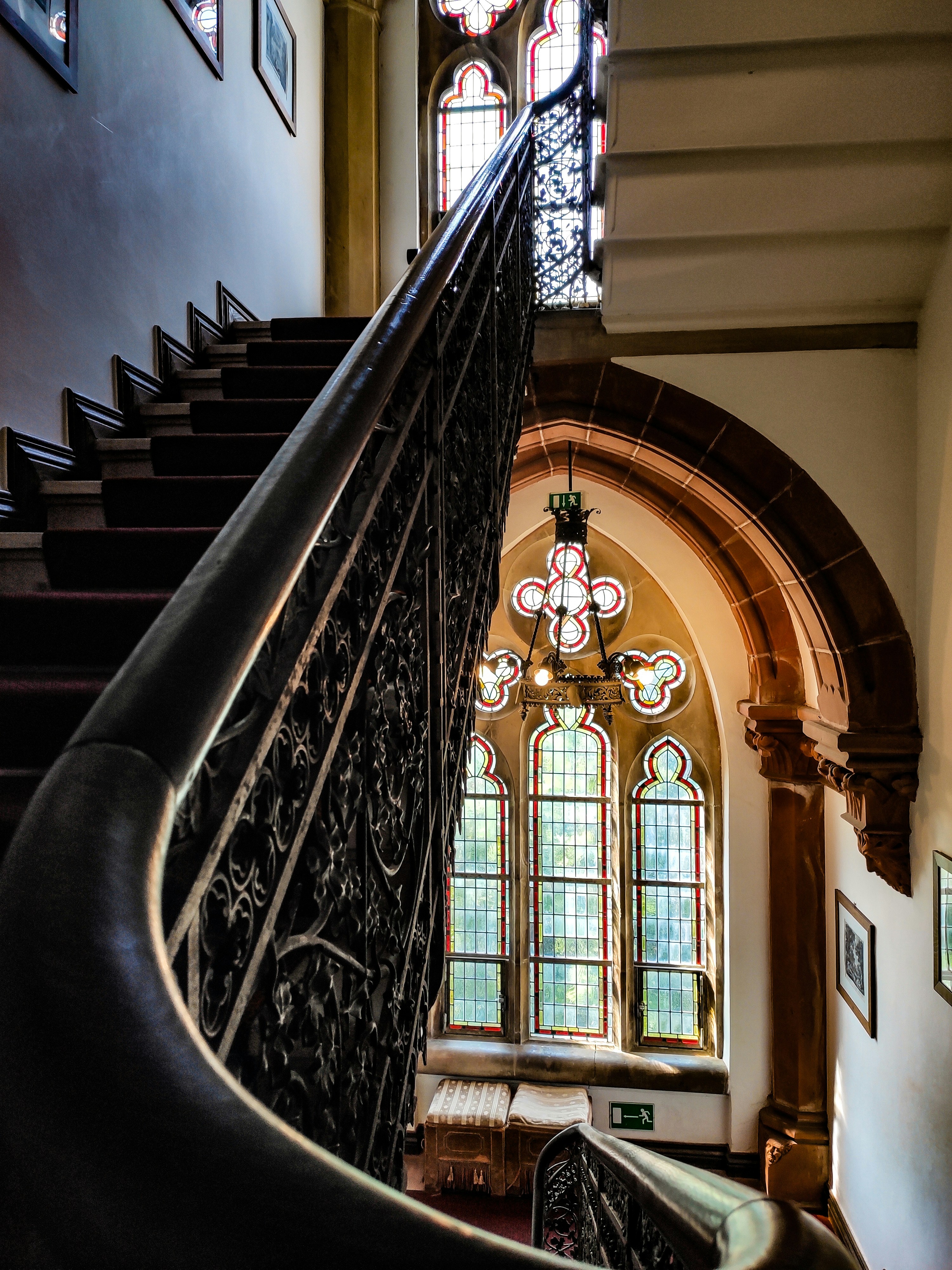 Intricate wrought iron railing leading up a staircase, illuminated by stained glass windows casting colorful light onto the walls.