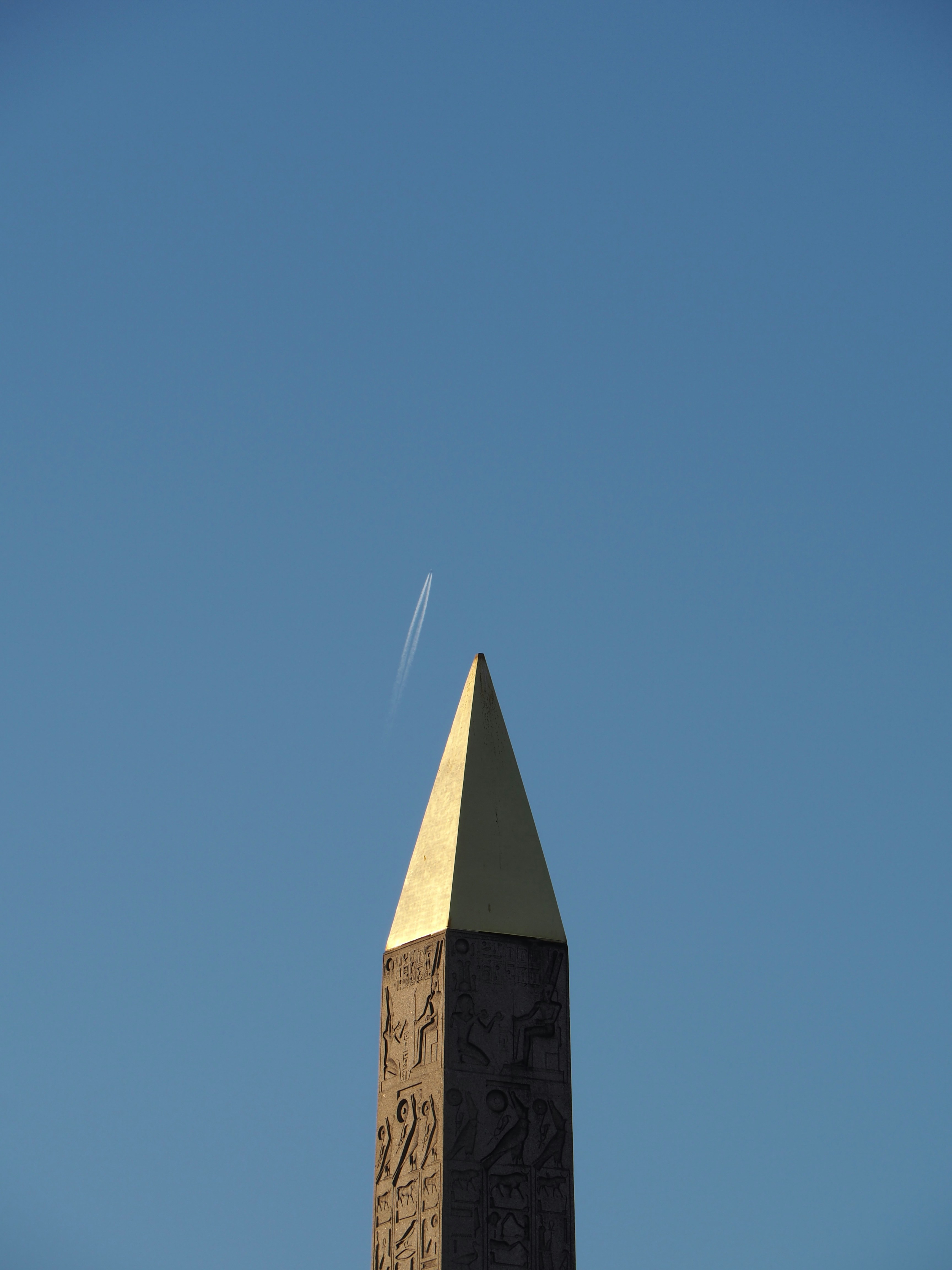 Tall obelisk with intricate carvings reaching towards a clear blue sky, crowned with a golden tip. A faint contrail traces the air above.