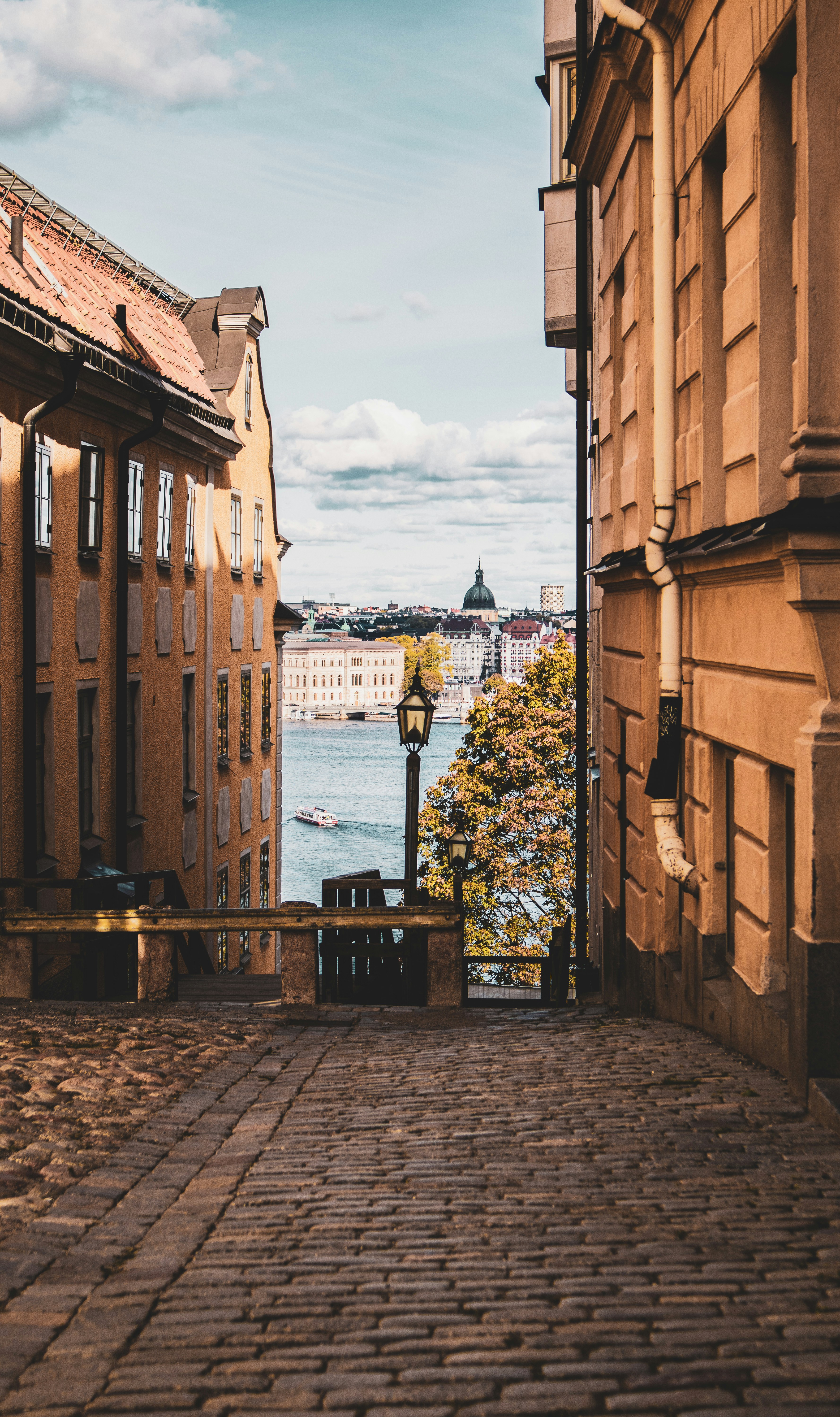 Charming cobblestone street framed by historic buildings leading to a tranquil waterfront view in Stockholm. A lamppost and autumn foliage add character to the scene.