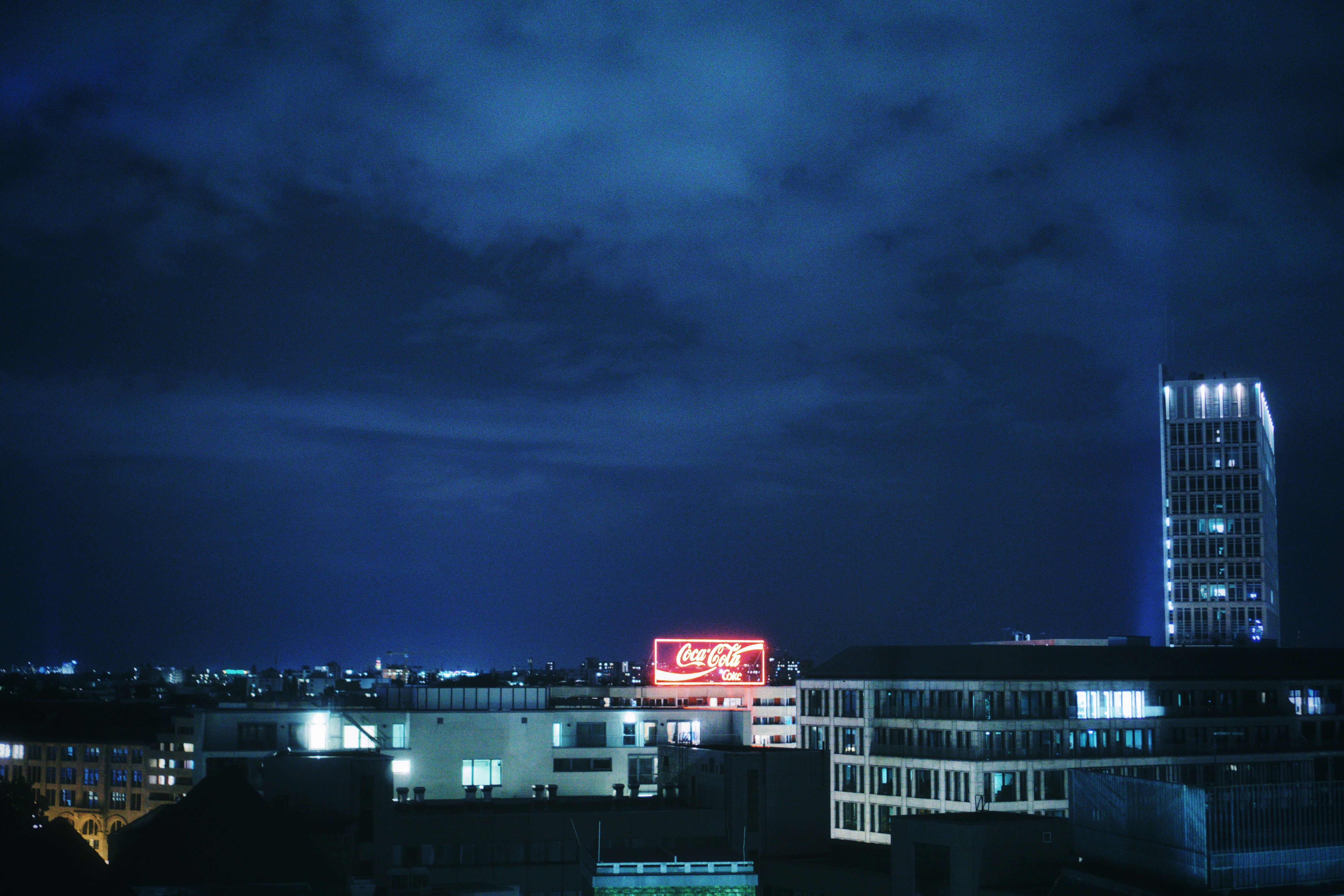 Illuminated Coca-Cola sign glows against a backdrop of city buildings under a moody night sky.