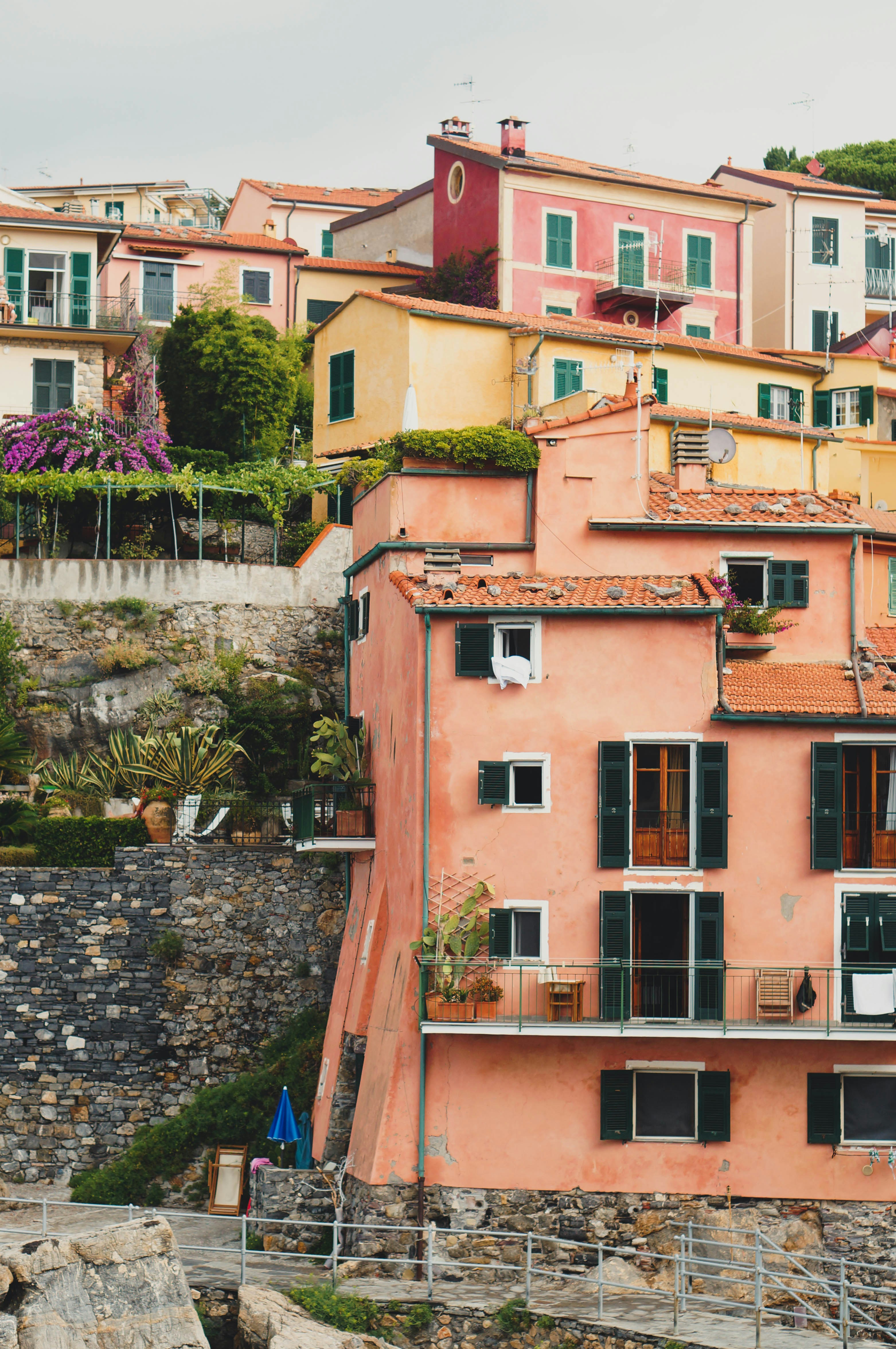 Pastel-colored hillside houses stack along a stone quay, their green shutters and terracotta roofs forming a vibrant coastal skyline. The scene captures a Mediterranean village perched on a cliff above the water.