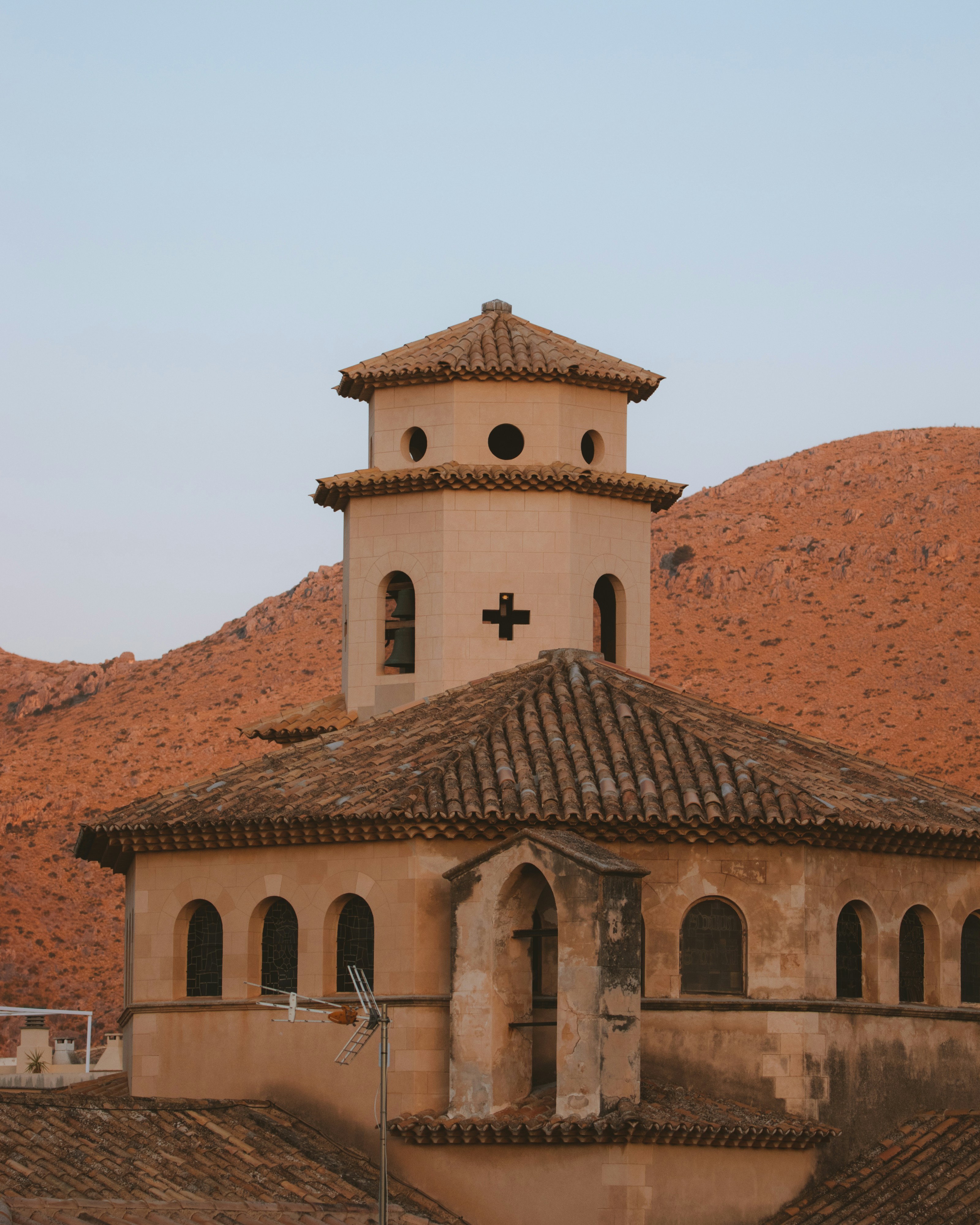 Historic building with a distinctive tower and cross, set against a backdrop of rugged mountains. The warm hues of the landscape enhance its architectural features.
