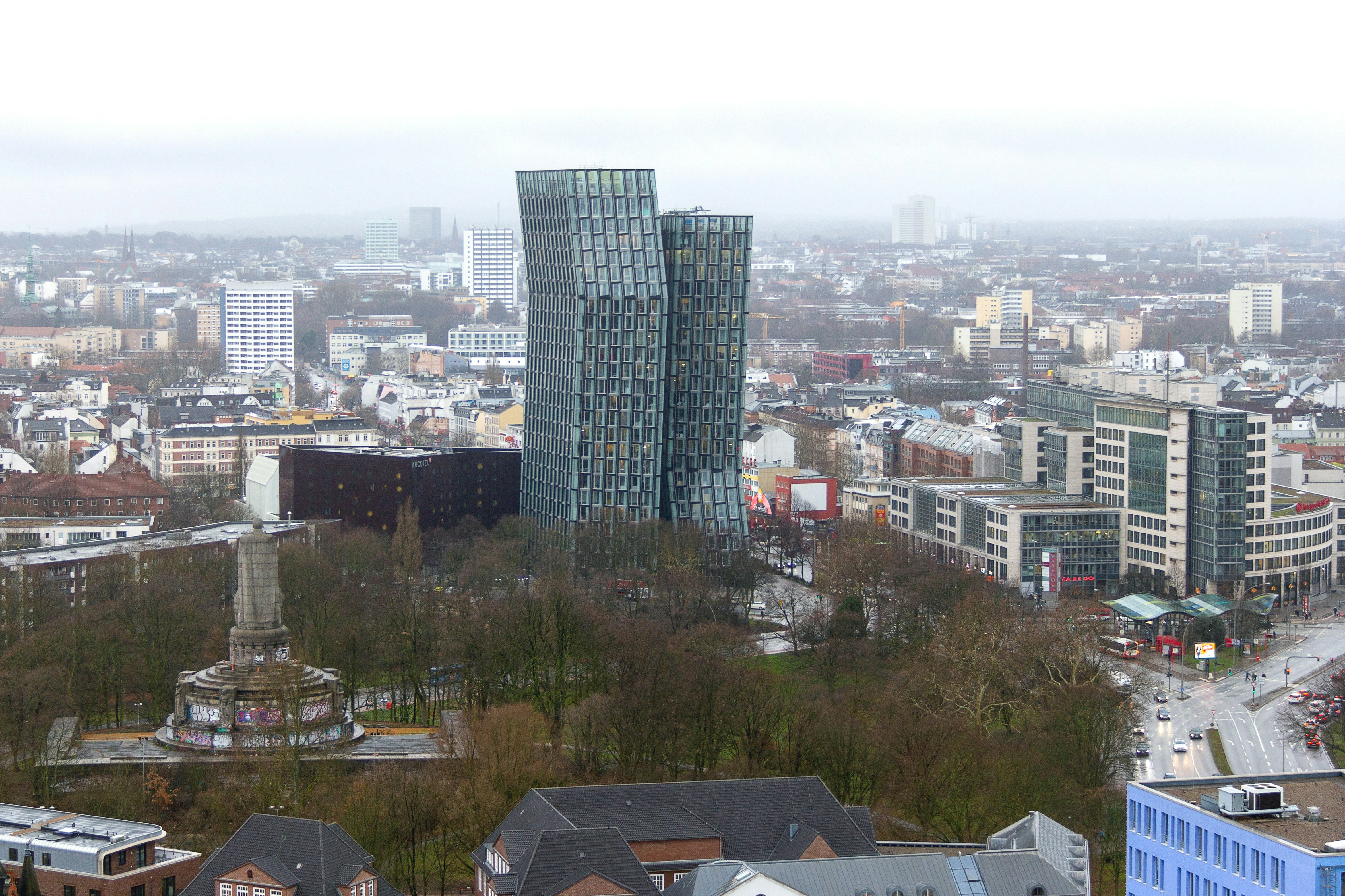 Modern glass building stands amidst a cityscape, surrounded by trees and historical structures, showcasing a blend of nature and urban life.