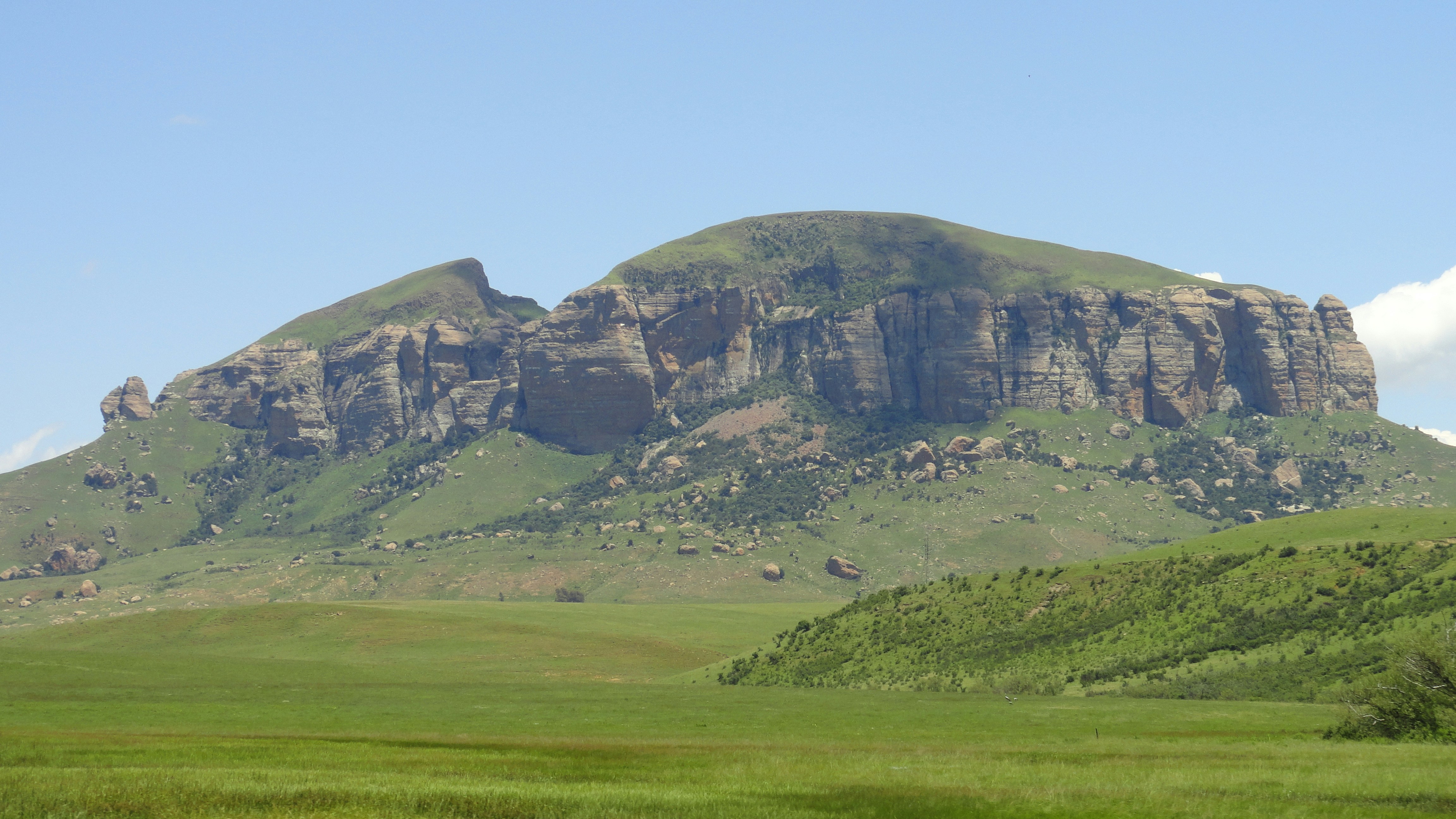 Rolling green plains lead to a rugged rock formation beneath a clear blue sky.
