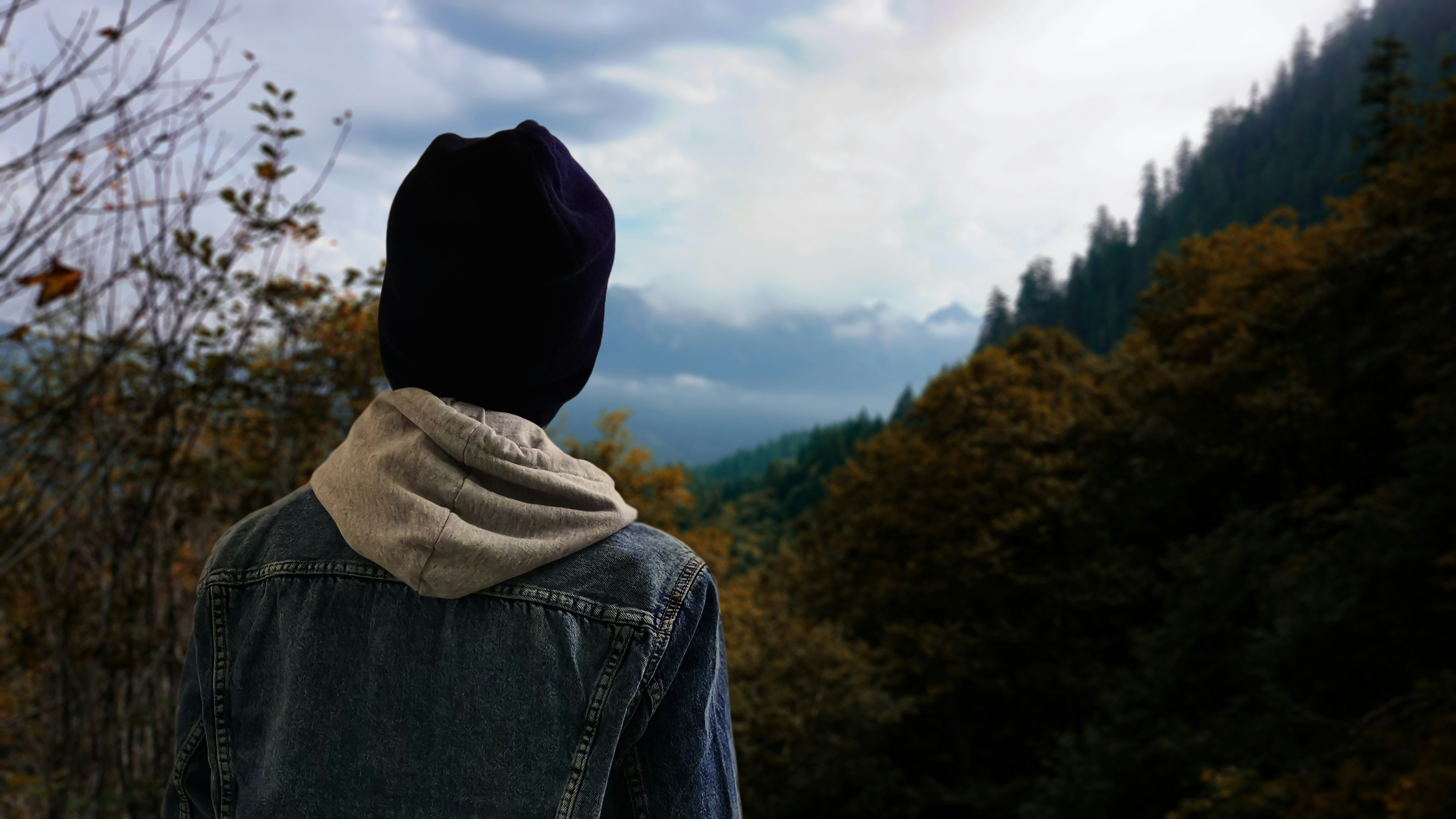 Person in a beanie and denim jacket gazing at a cloudy mountain landscape.