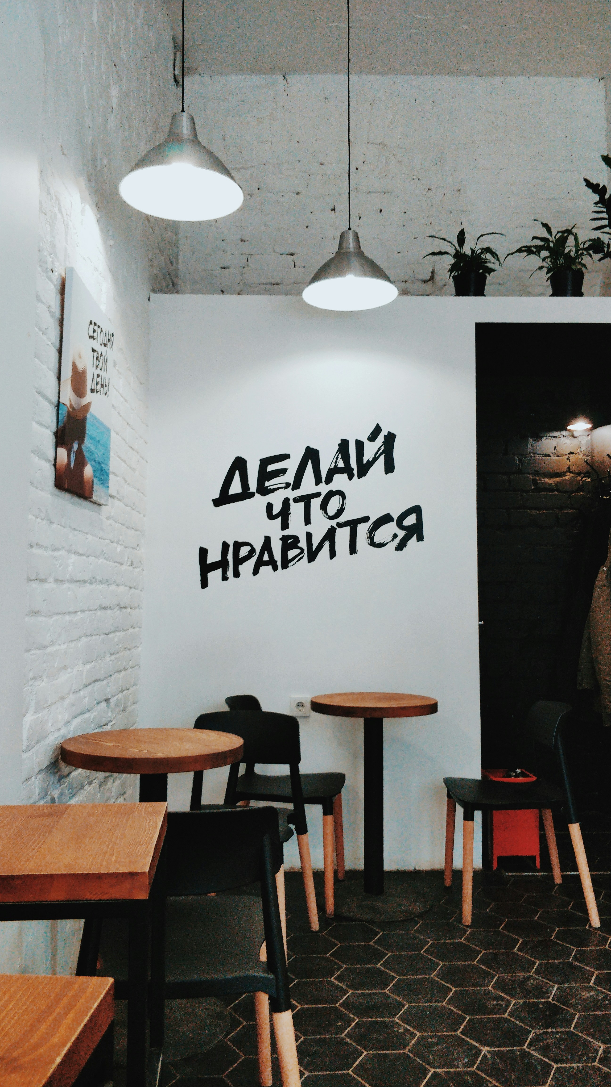 Stylish café interior featuring wooden tables, black chairs, and a motivational phrase on the wall. The ambiance is enhanced by hanging lights and greenery.