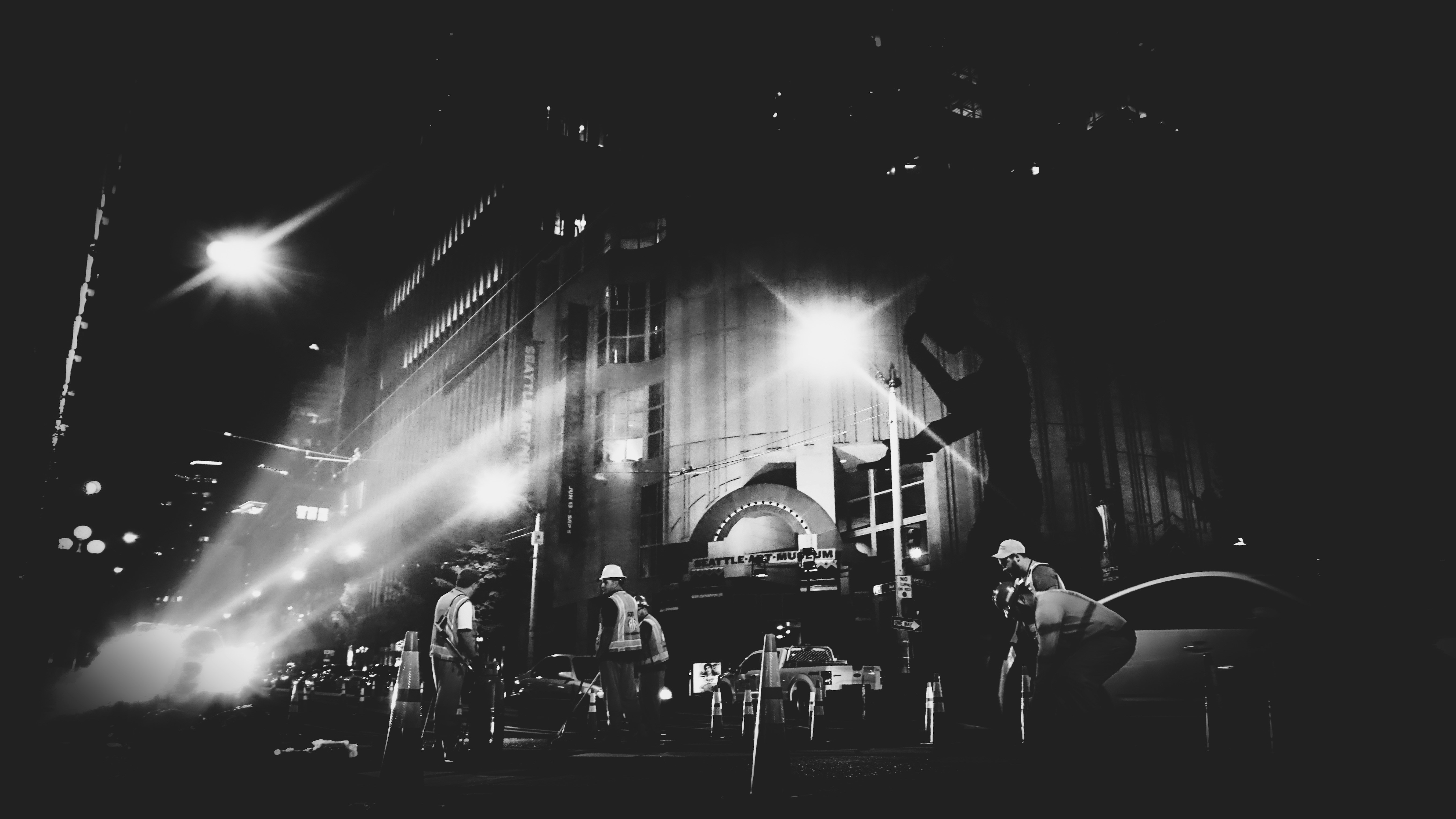 Construction workers engaged in nighttime activity amidst a bustling cityscape, illuminated by streetlights and building facades.