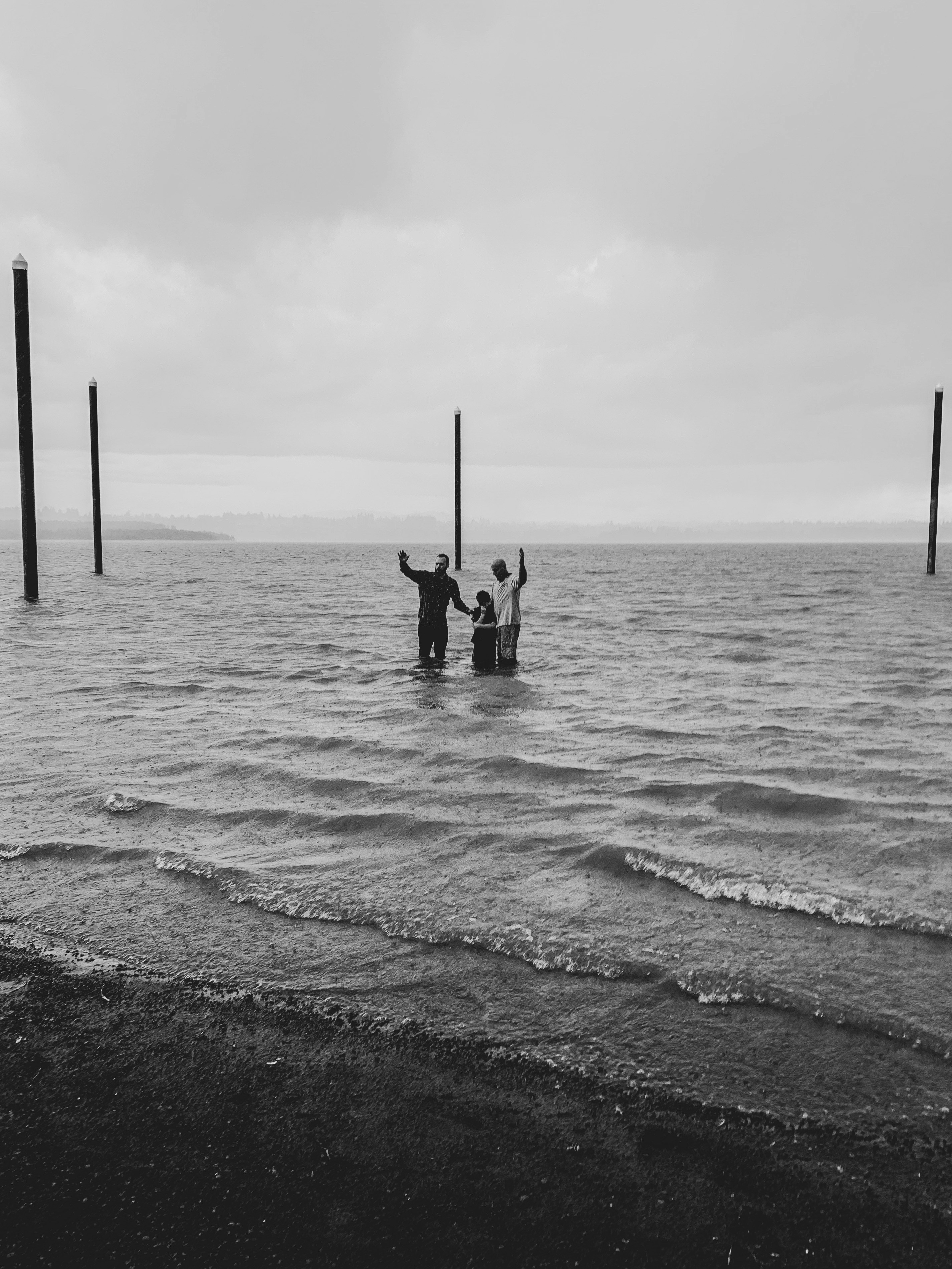 Two figures wade through a flooded area, silhouetted against a backdrop of vertical poles and choppy waters. The scene captures a moment of resilience amidst nature's challenges.