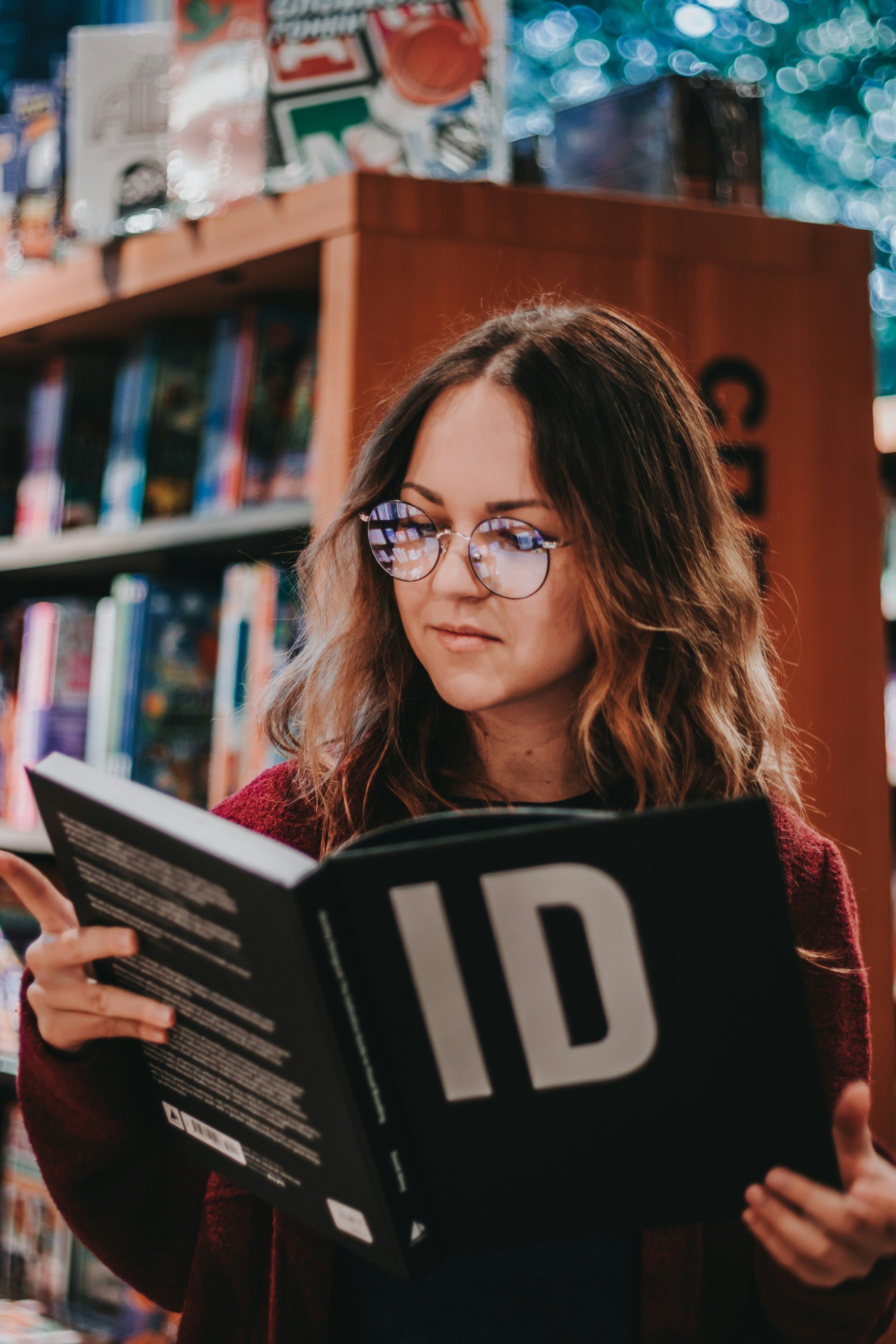 A woman with round glasses reads a bold black book titled 'ID' in a colorful bookstore, with shelves and soft neon bokeh in the background.