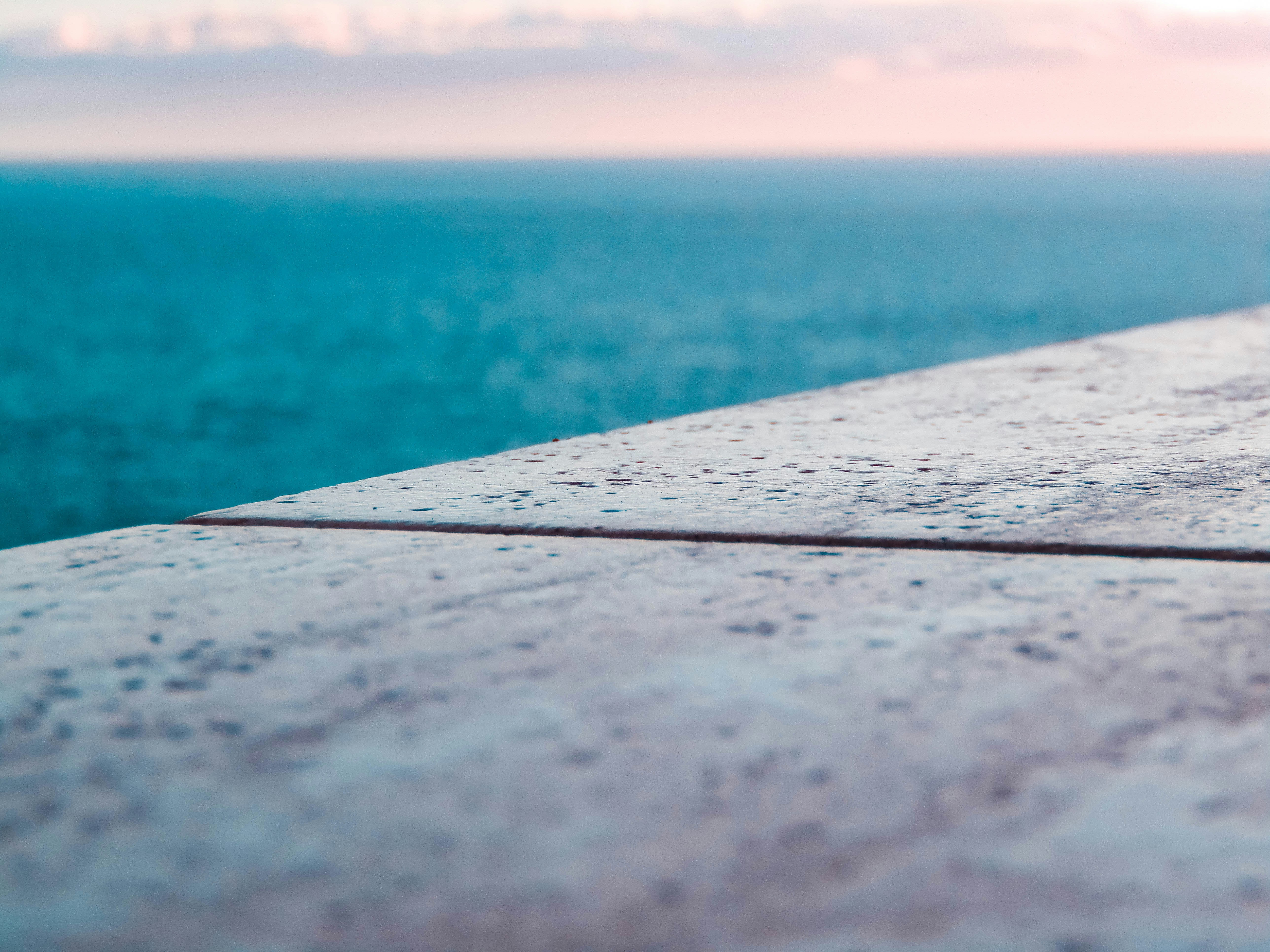 Shallow-focus photograph of a weathered deck edge leading toward a calm sea at sunset, with a pastel sky.