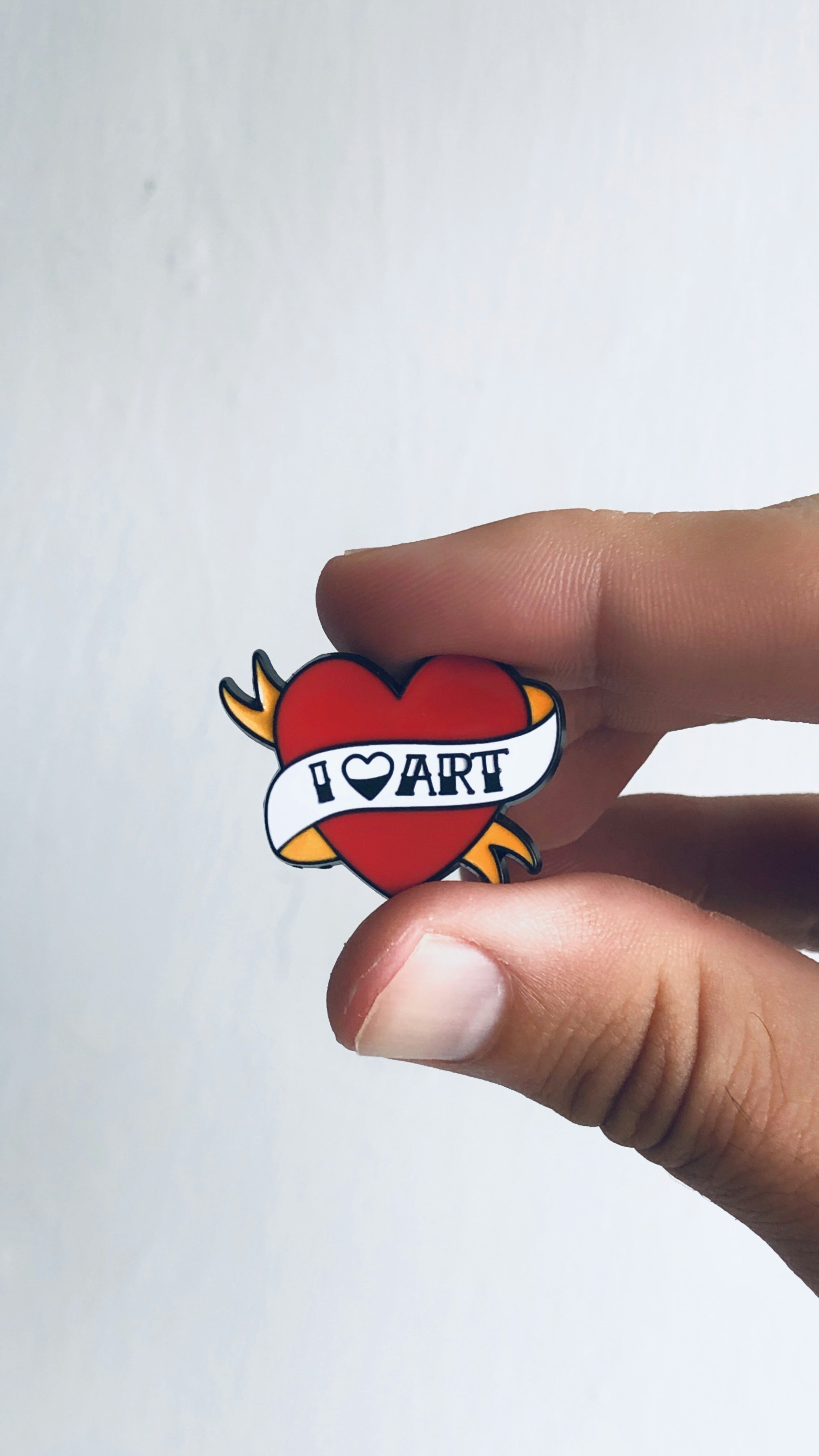 A hand holding a vibrant red heart pin adorned with a banner that reads 'I ♥ ART'. The background is softly blurred, emphasizing the pin's details.