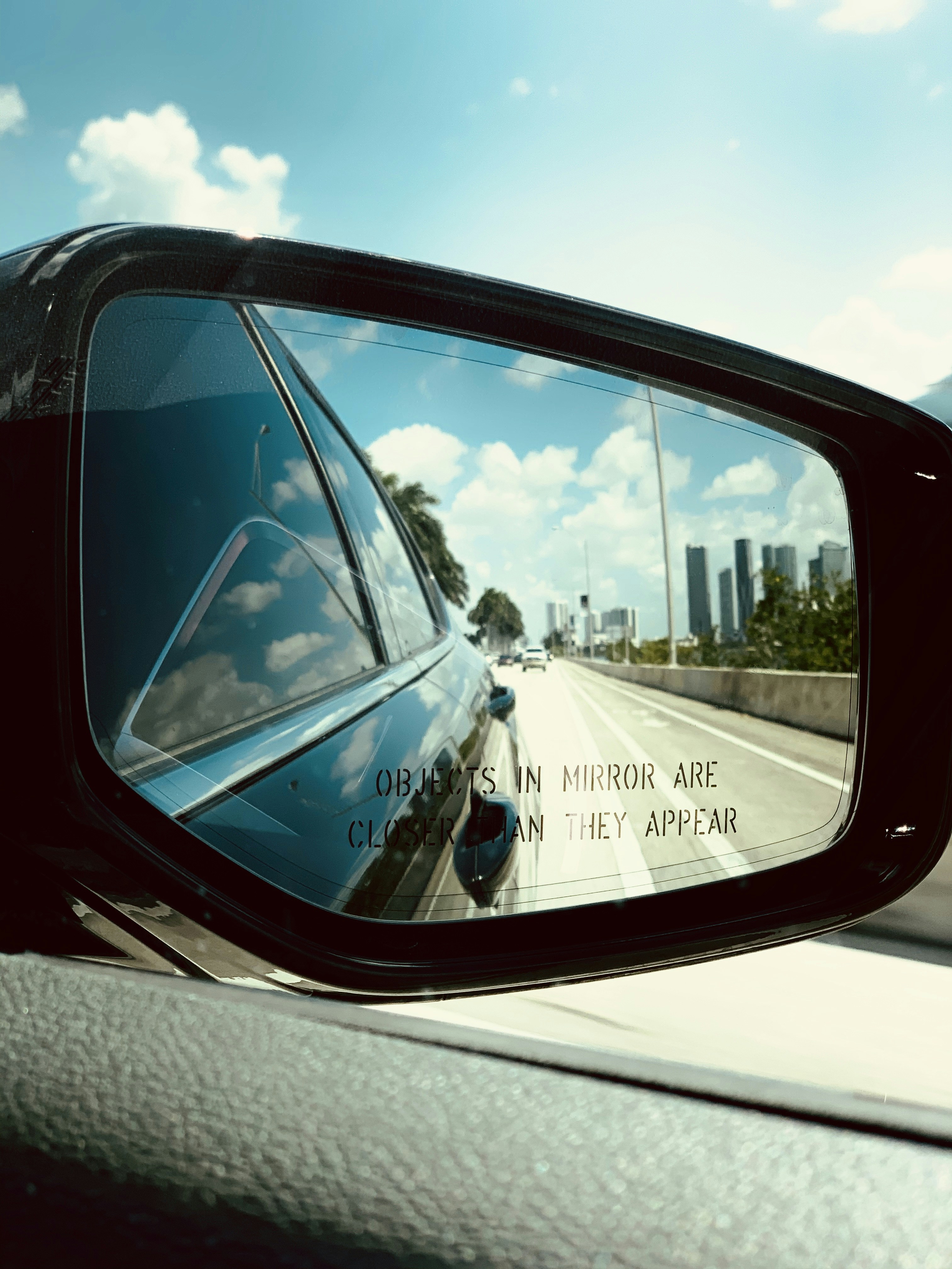Side mirror reflecting a cityscape with clouds above, featuring the warning 'Objects in mirror are closer than they appear.'