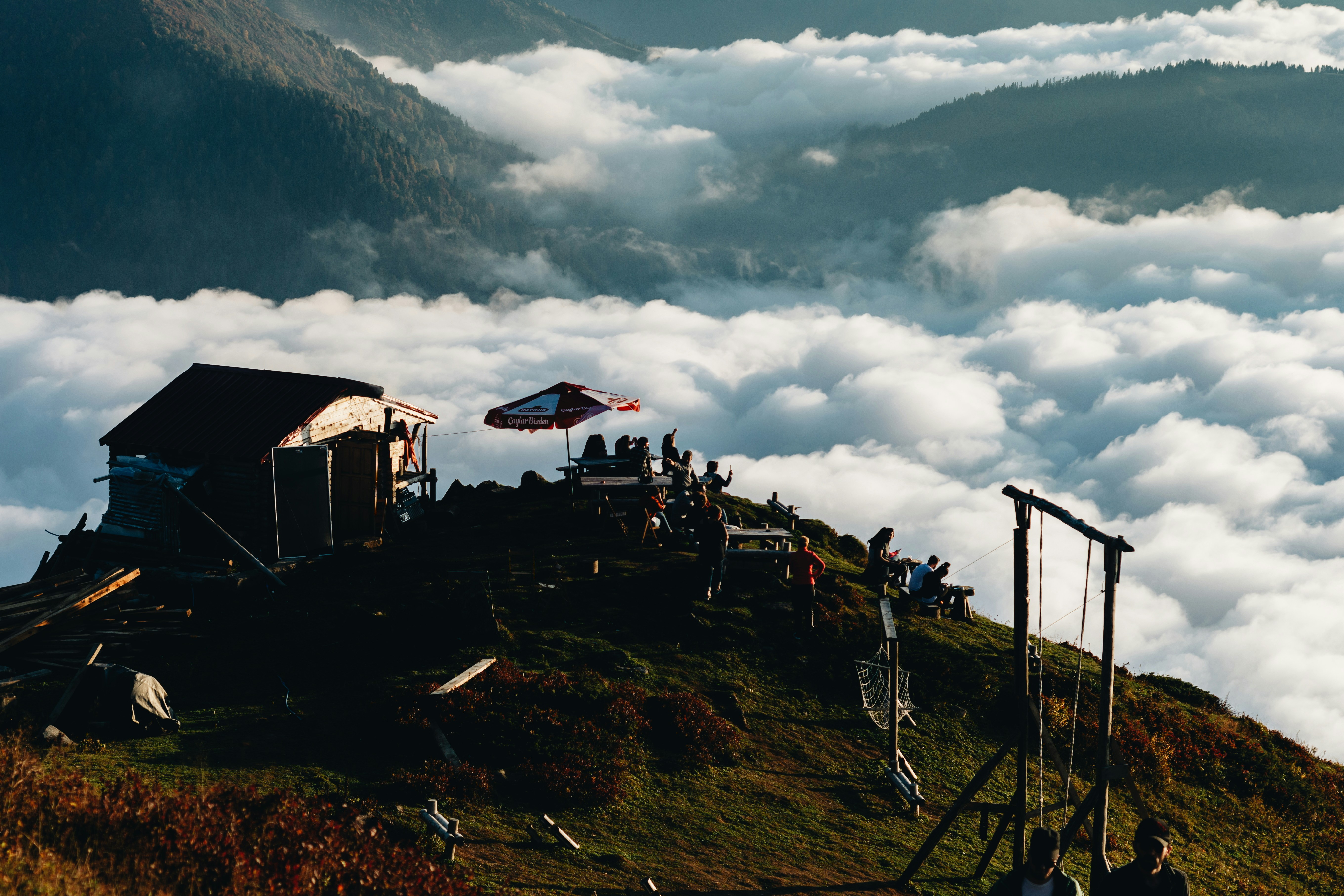 A rustic cabin overlooking a sea of clouds with people enjoying the view, set against a backdrop of mountains. The scene conveys a sense of tranquility and connection with nature.