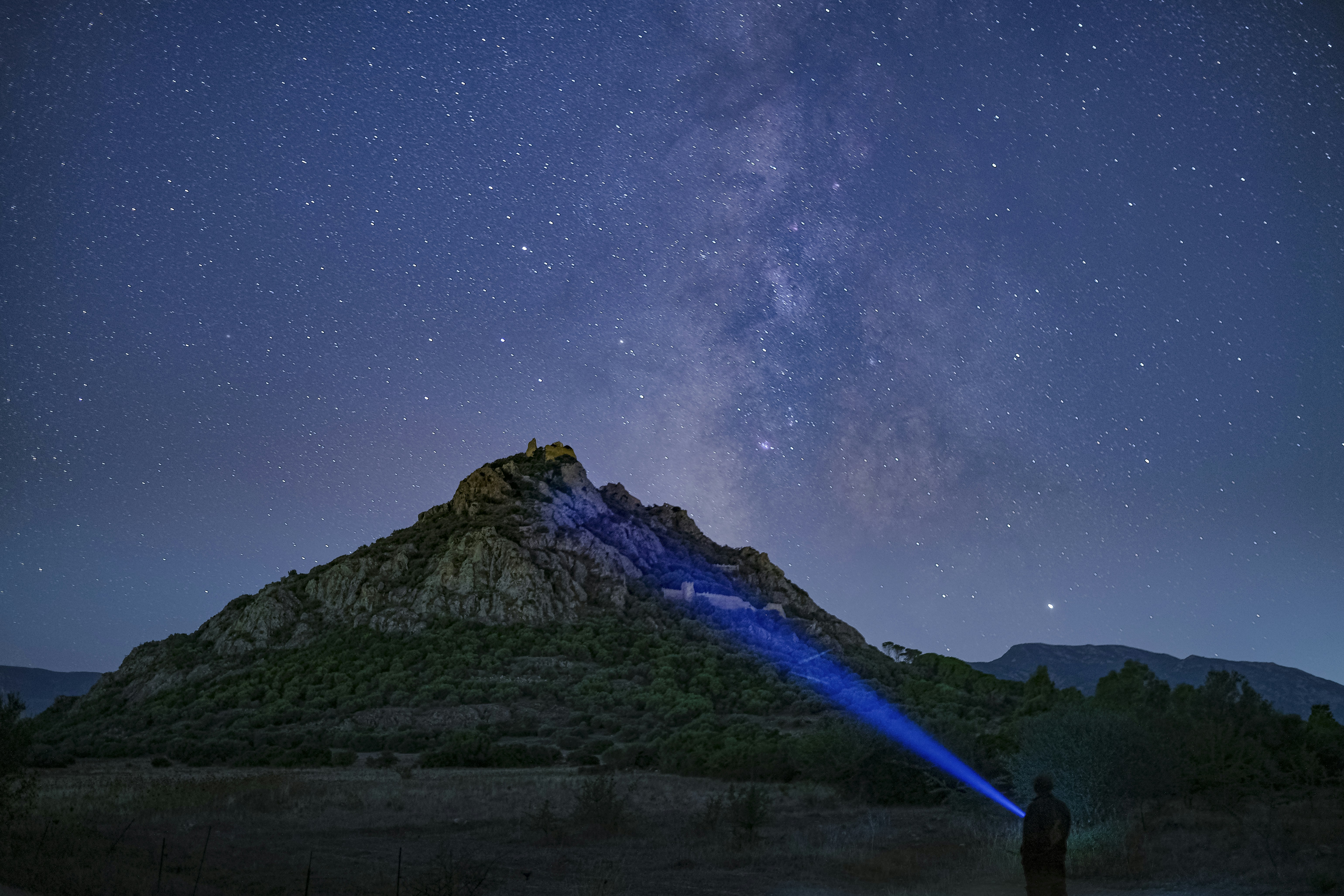 A figure stands at the base of a rocky mountain, casting a blue beam into the starry night sky, revealing the Milky Way's splendor.
