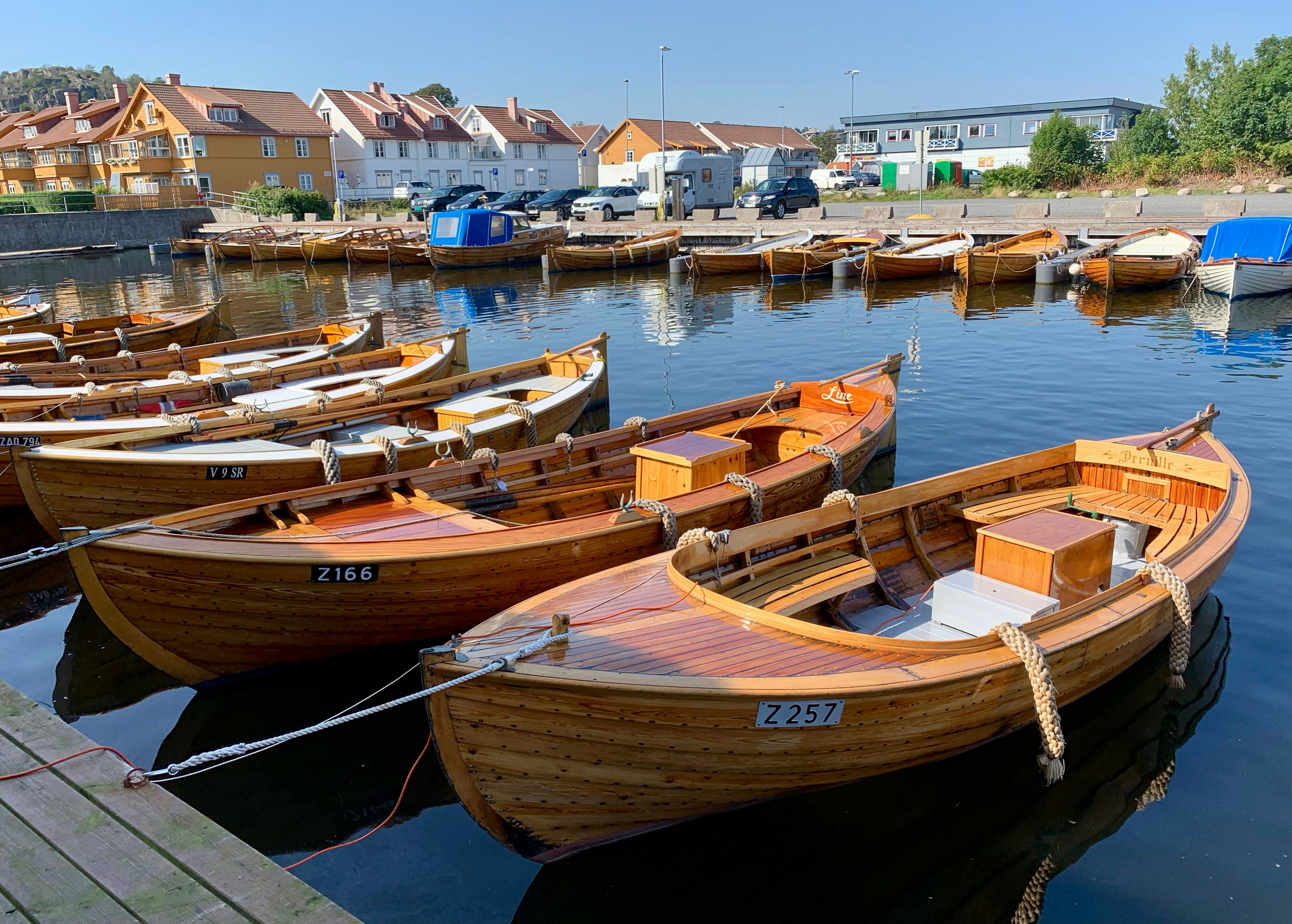 Rowboats moored in a tranquil marina, showcasing their polished wooden finishes against the calm waters. The backdrop features charming coastal buildings.