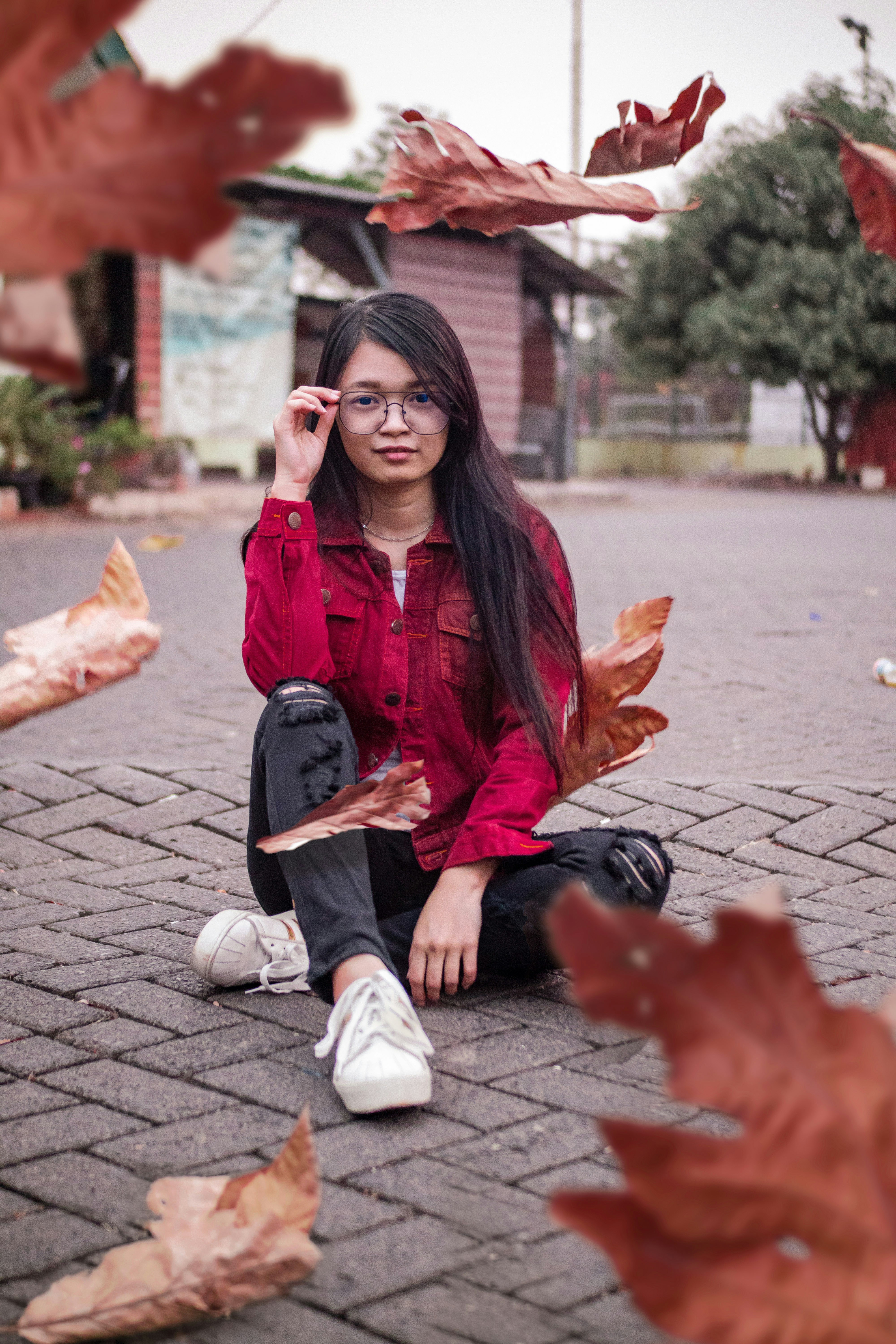 A young woman in a red jacket poses amidst swirling autumn leaves on a cobblestone path, evoking a sense of seasonal change.