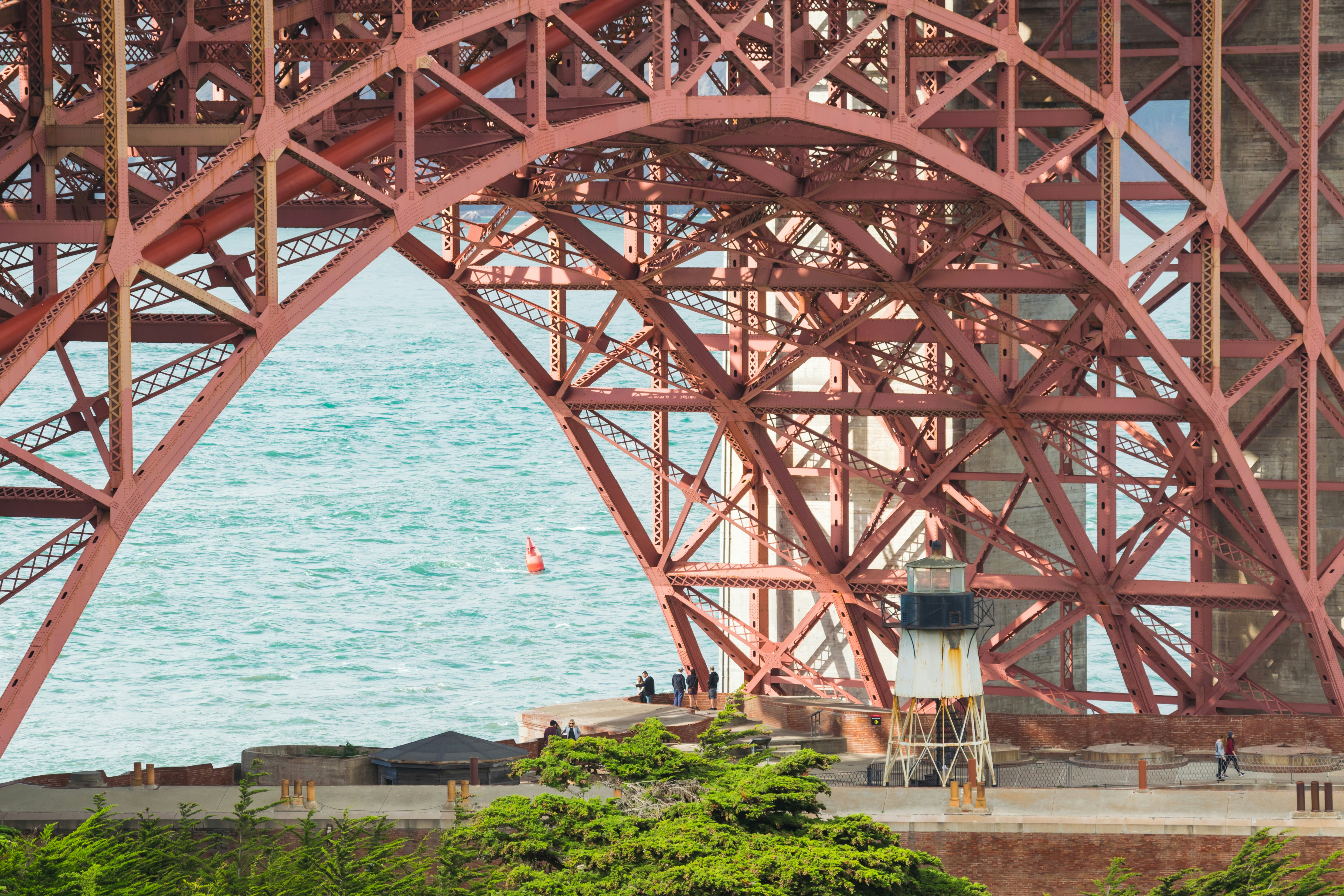 Intricate metalwork of a bridge's understructure frames a serene coastal scene, featuring a lighthouse and figures along the shore.