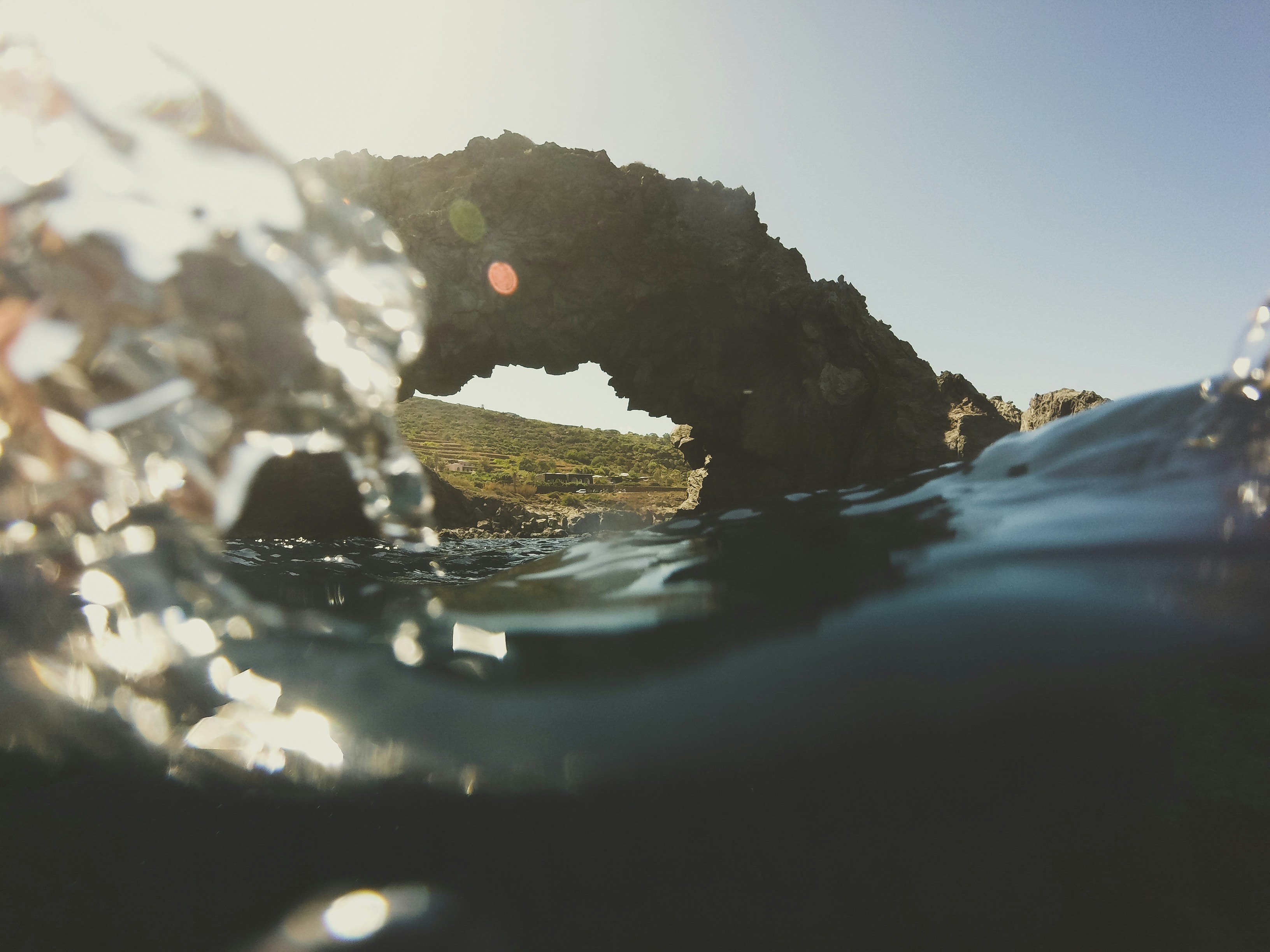 Ocean water splashes near a rocky arch under the bright sun.