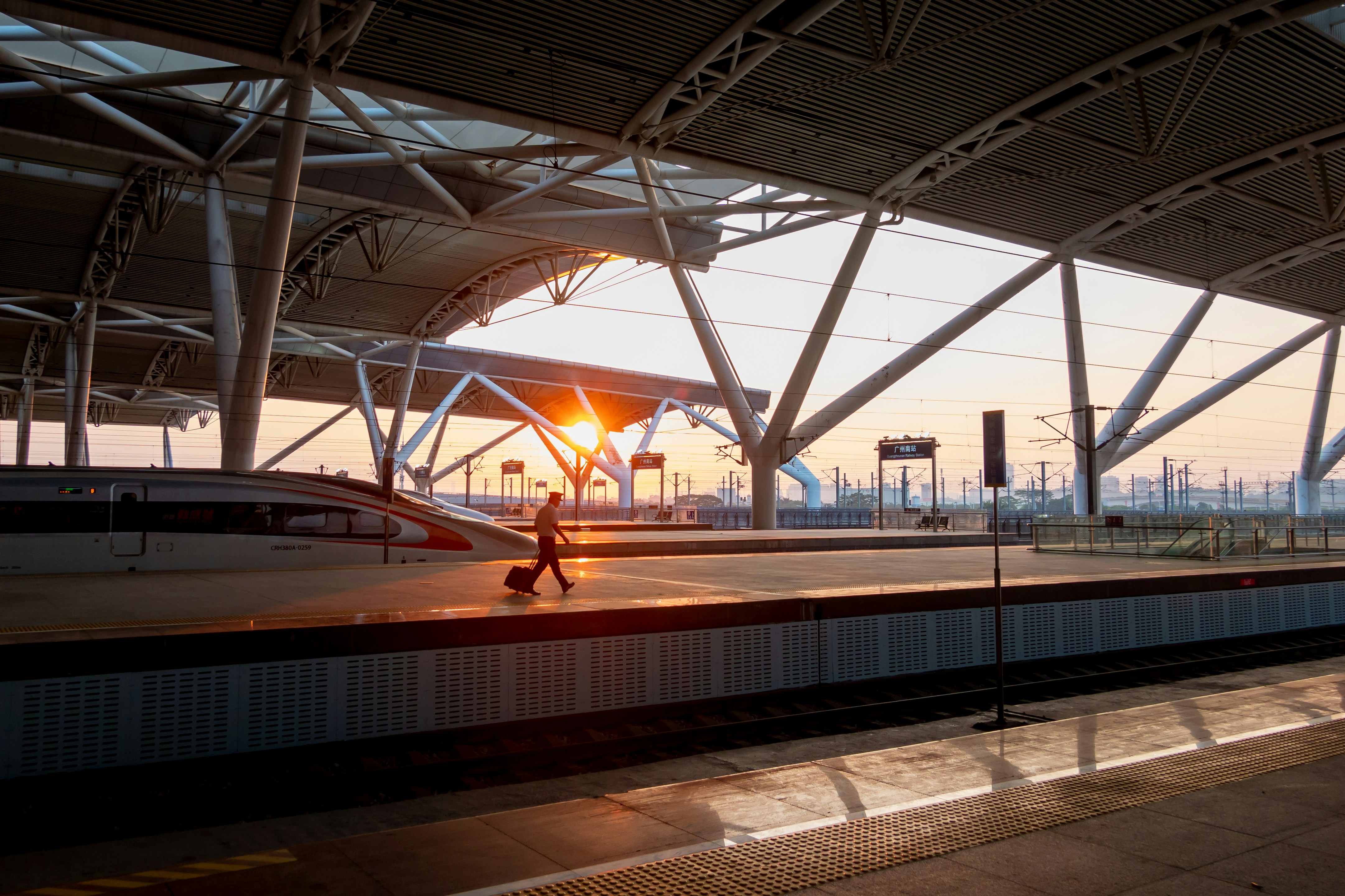 Traveler with a suitcase walks on an empty train platform at sunrise, framed by modern architectural beams.