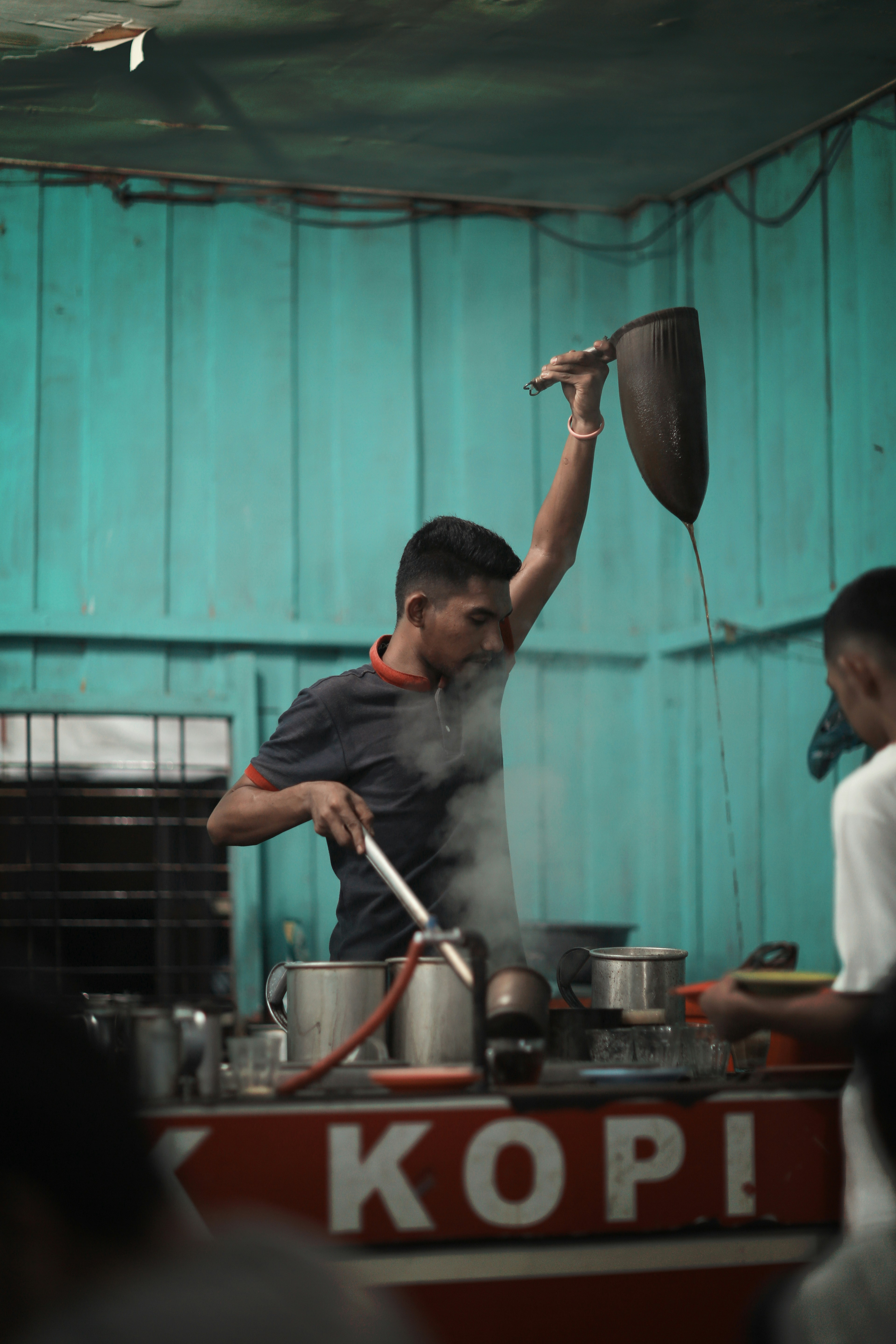 Barista pouring coffee from a traditional vessel, surrounded by steaming pots in a vibrant café setting.