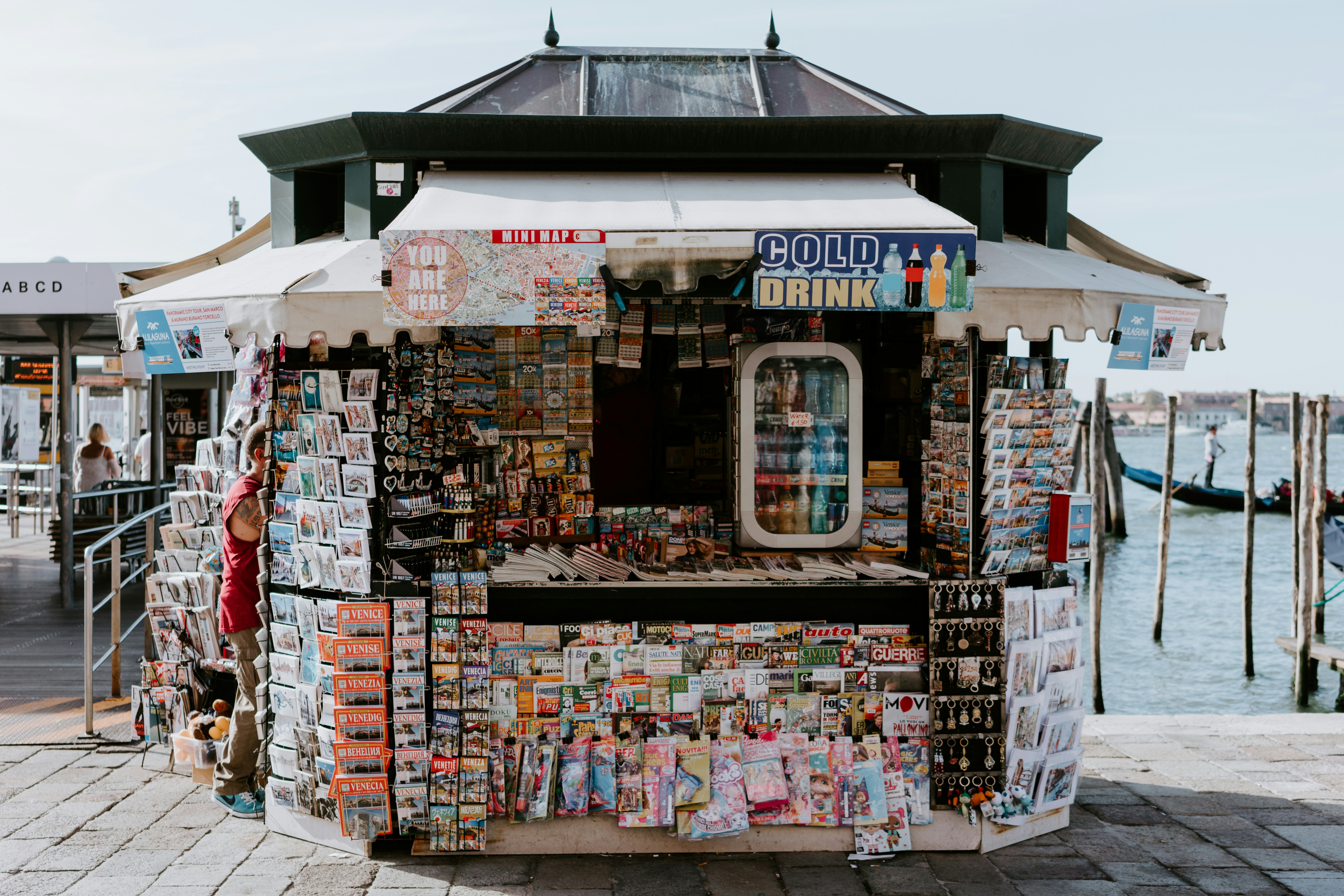 A bustling kiosk adorned with colorful magazines and drinks, set against a serene waterfront backdrop. The lively atmosphere invites passersby to explore its offerings.