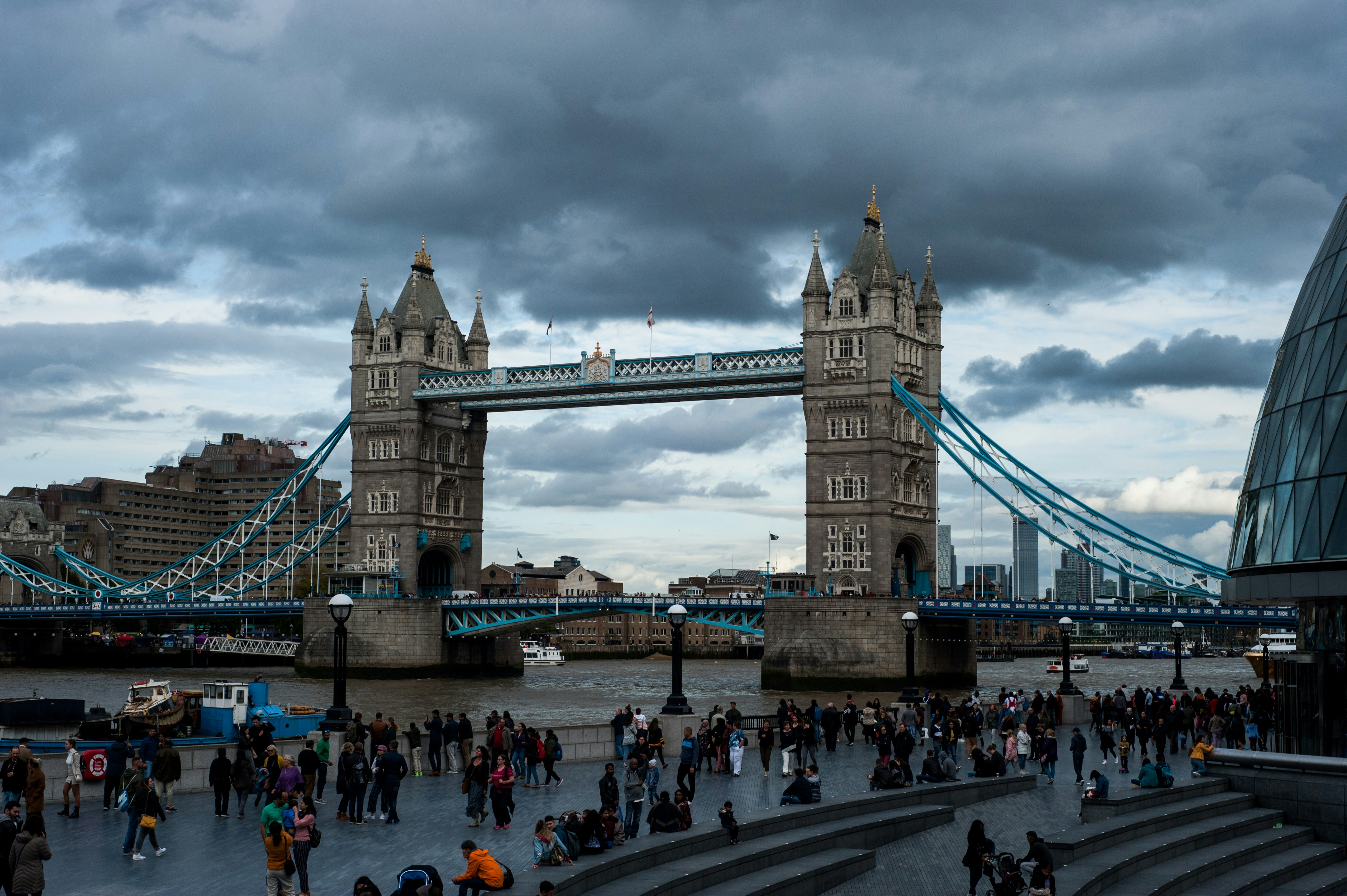 Tower Bridge spans the River Thames, flanked by a bustling crowd and dramatic clouds overhead.