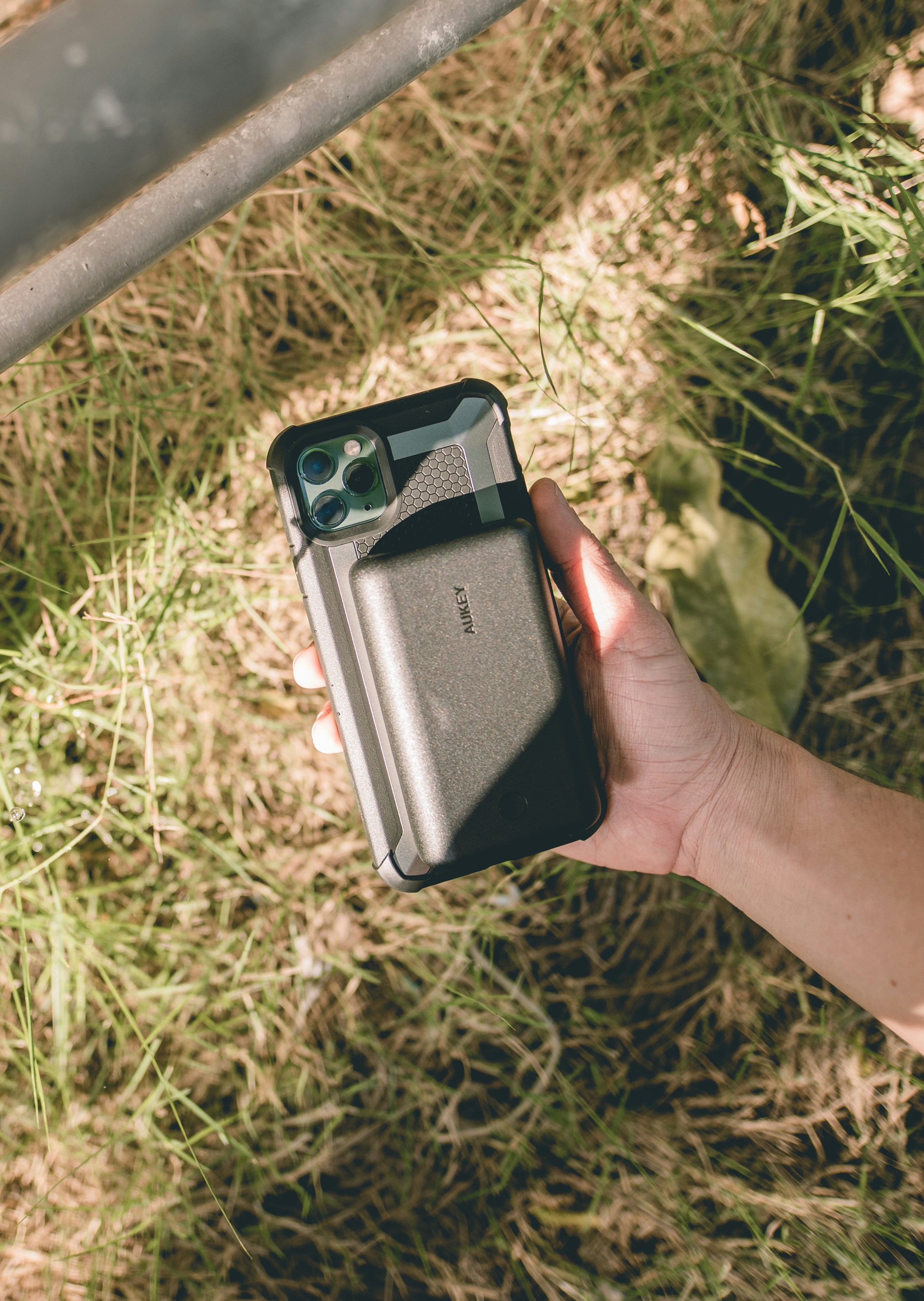 A hand holds a smartphone with a sleek battery pack against a backdrop of grass and foliage. The scene highlights the fusion of technology and nature.