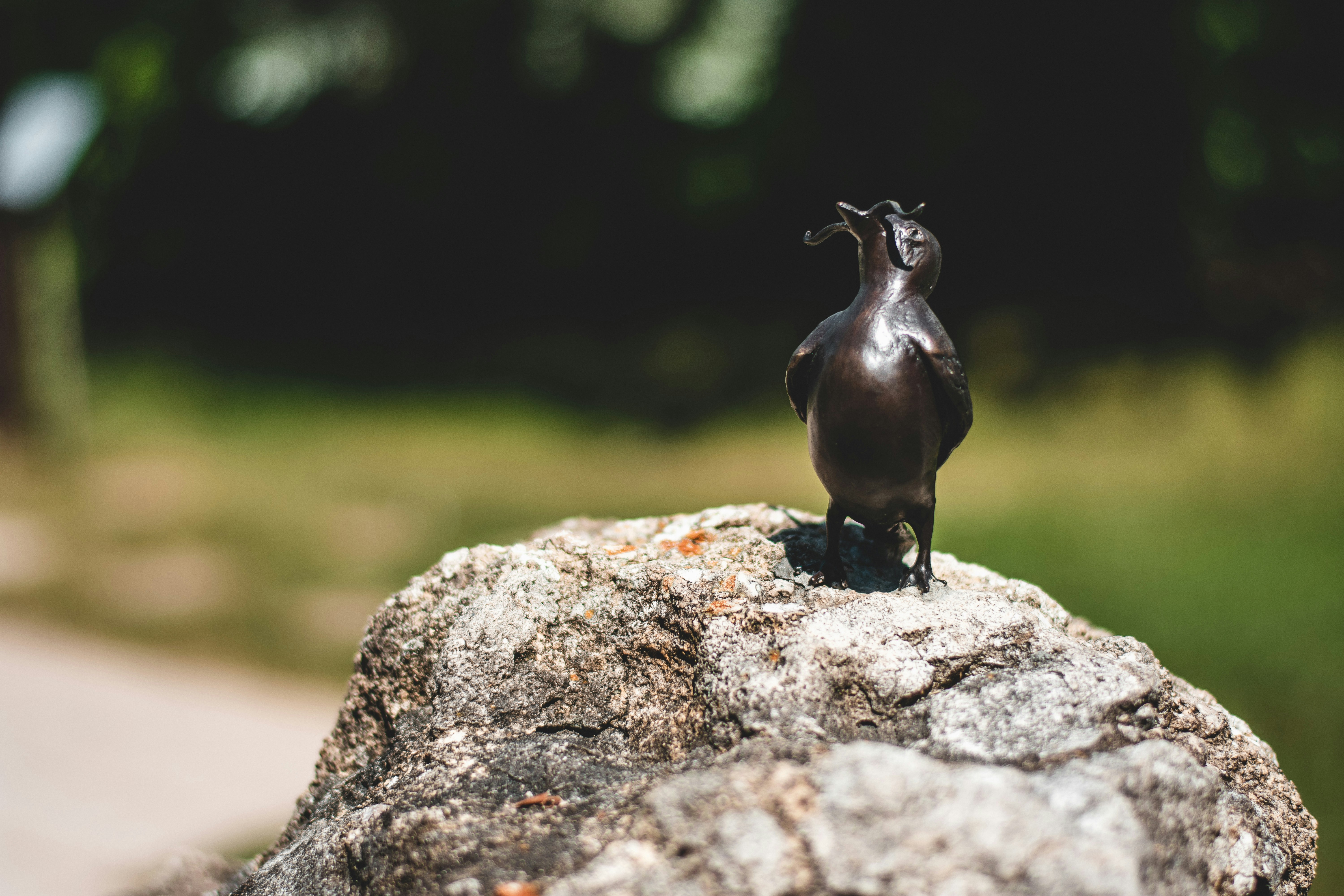 Bronze bird sculpture perched on a rock, gazing into the distance amidst a blurred natural backdrop. 