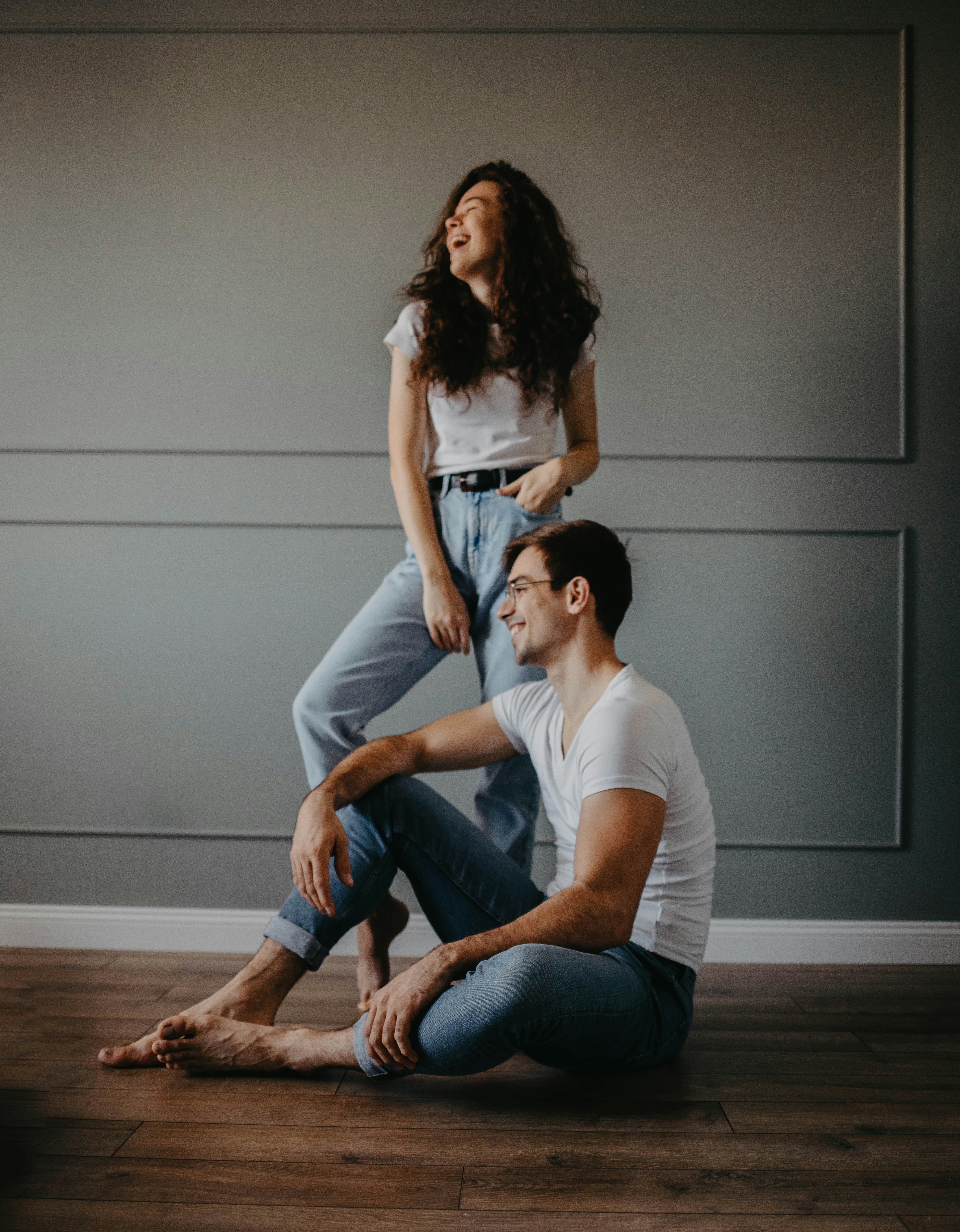 Two individuals share a light-hearted moment in a stylish indoor setting, showcasing relaxed fashion against a muted backdrop.