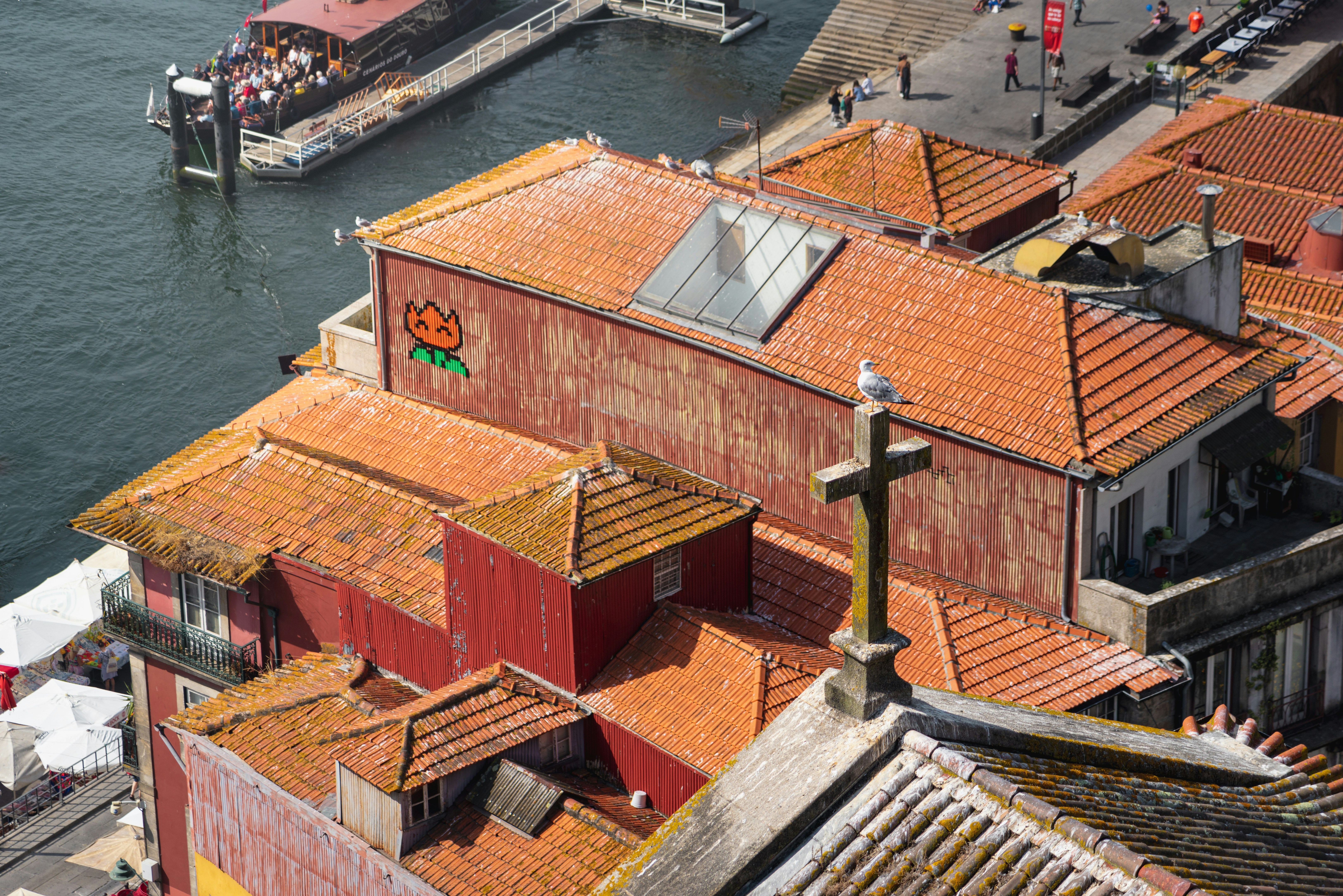 Vibrant rooftops adorned with orange tiles and a playful mural overlook a bustling waterfront scene. A cross stands prominently, adding a touch of history to the lively urban landscape.