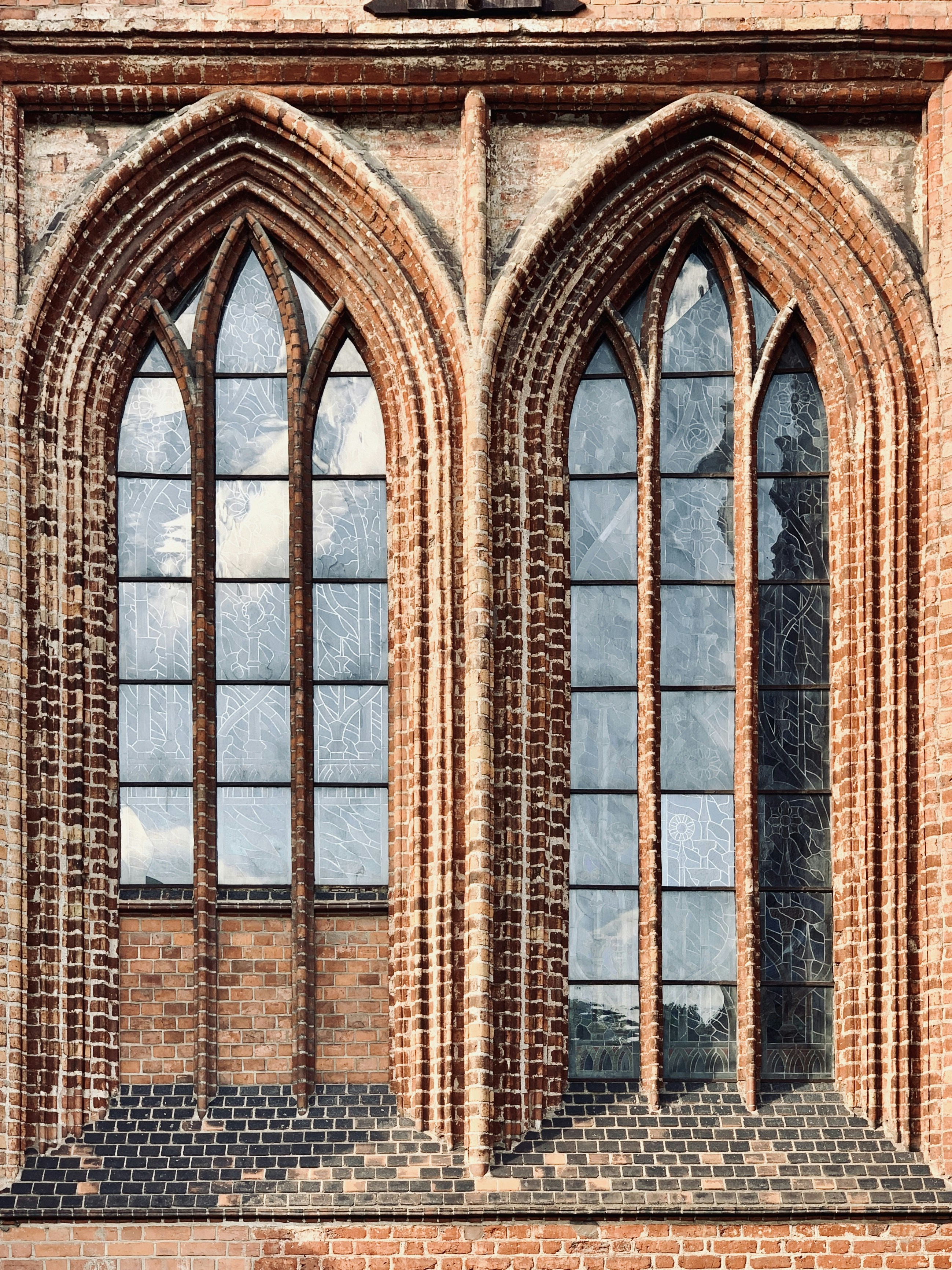 Intricate Gothic-style windows framed by textured brickwork, reflecting the sky and surroundings. The composition highlights the interplay of light and structure.