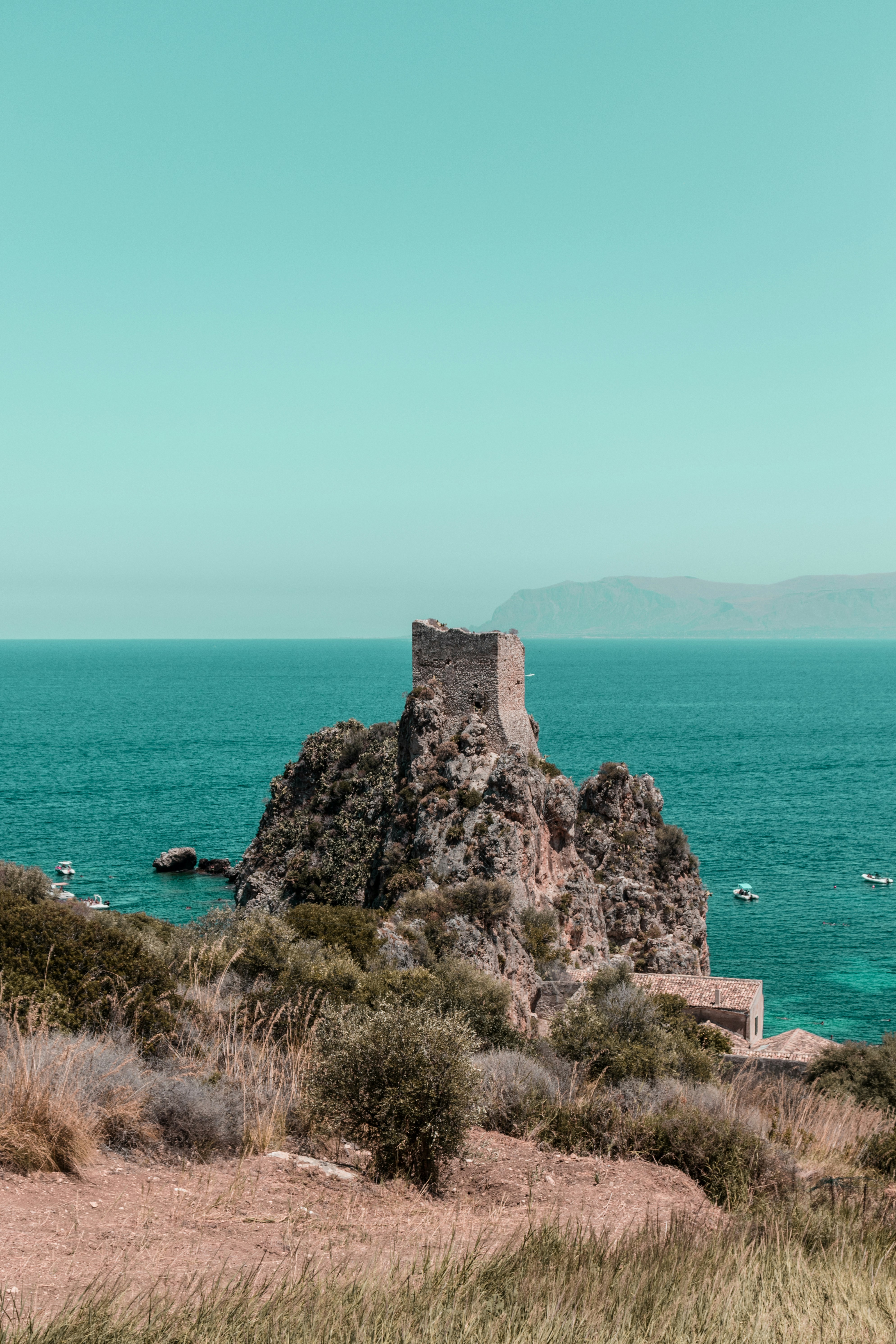 Ancient stone tower perched on a rocky outcrop overlooking a serene turquoise sea, surrounded by lush greenery.
