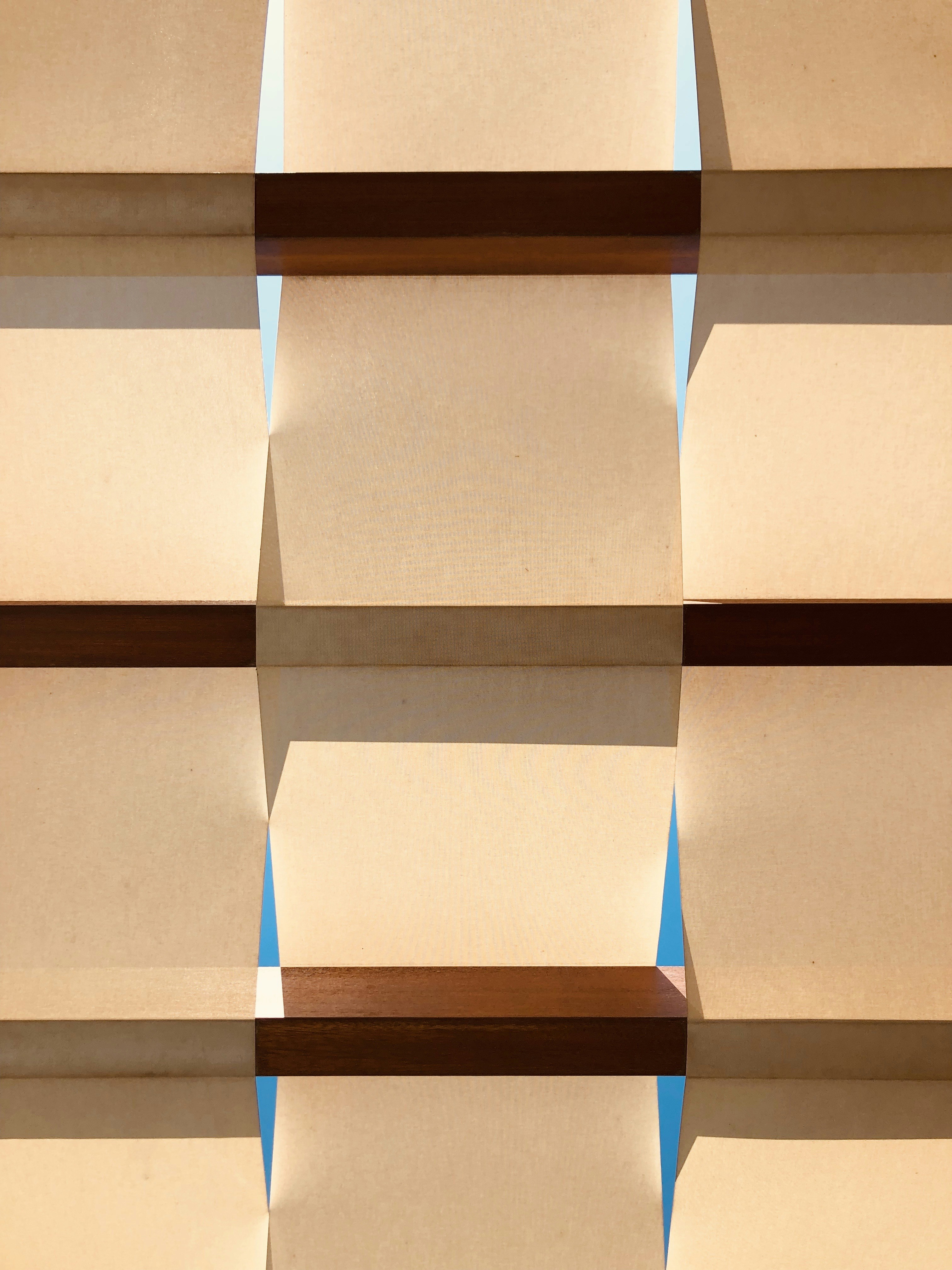 Layered paper and wooden slats create a rhythmic pattern, filtering sunlight and casting soft shadows. The blue sky peeks through, enhancing the contrast.