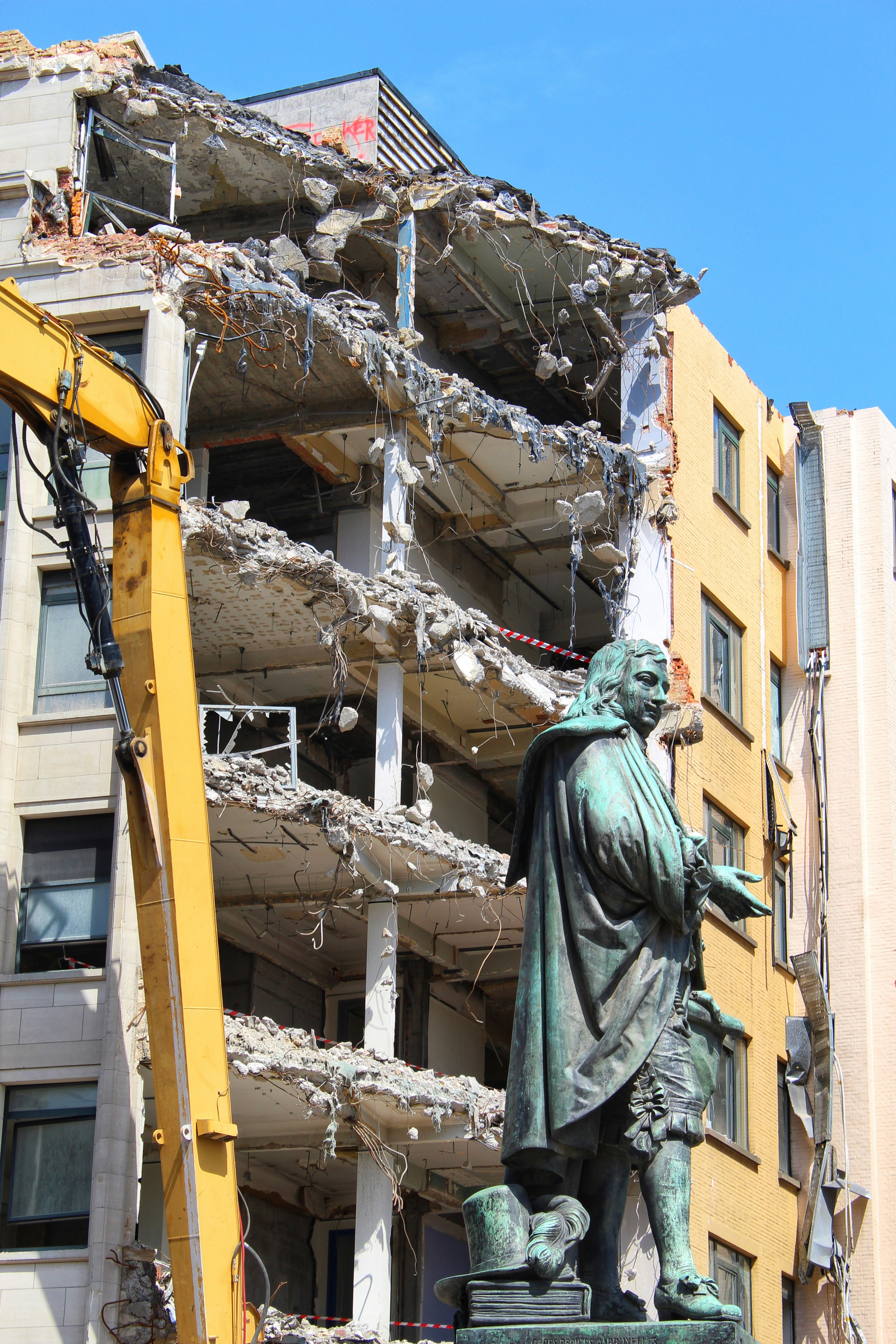 Bronze Guardian at the Edge of Collapse: Statue Fronting a Damaged Facade and a Yellow Excavator