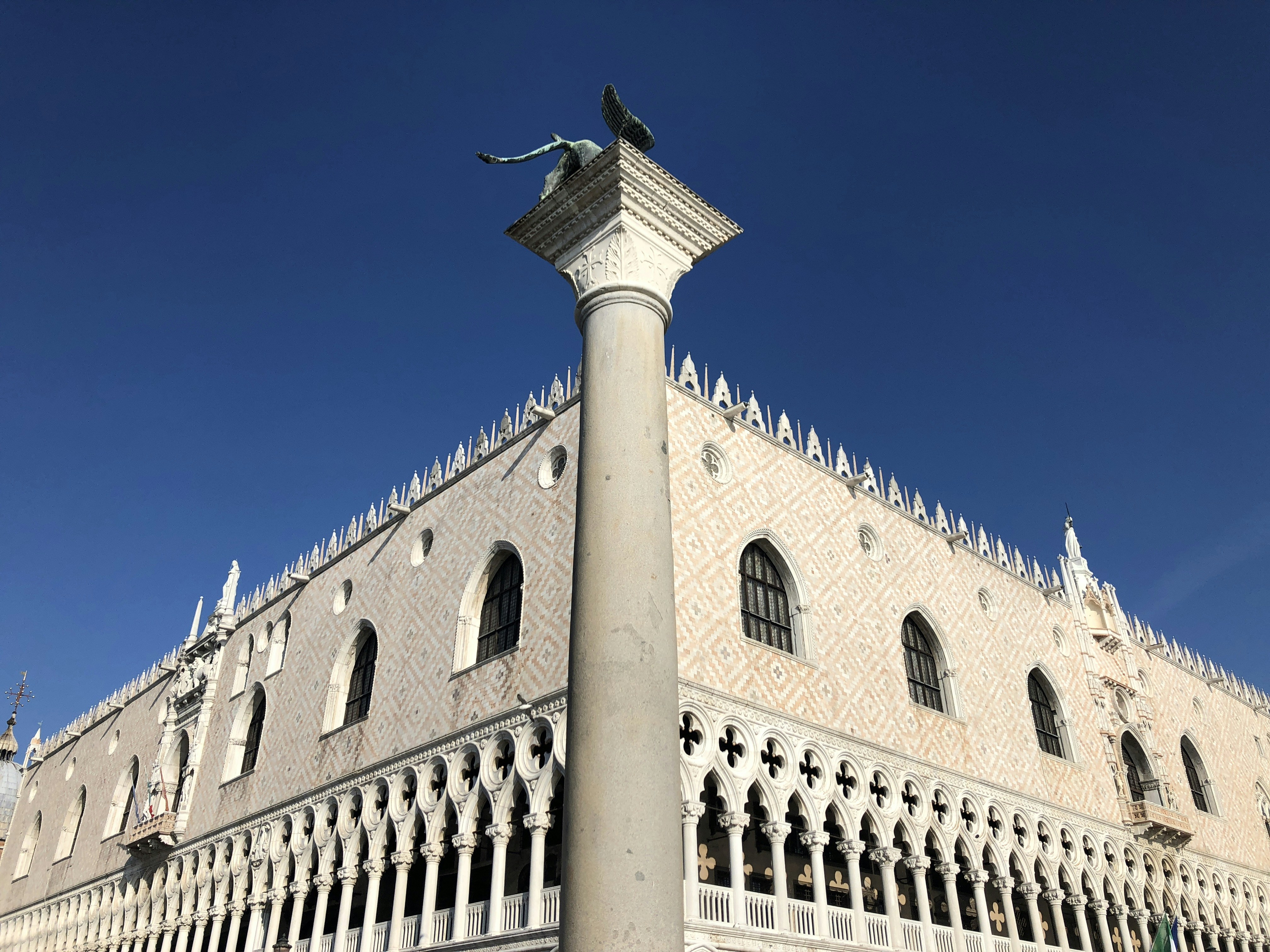 Column topped with a statue stands before the ornate façade of a Venetian palace under a clear blue sky.