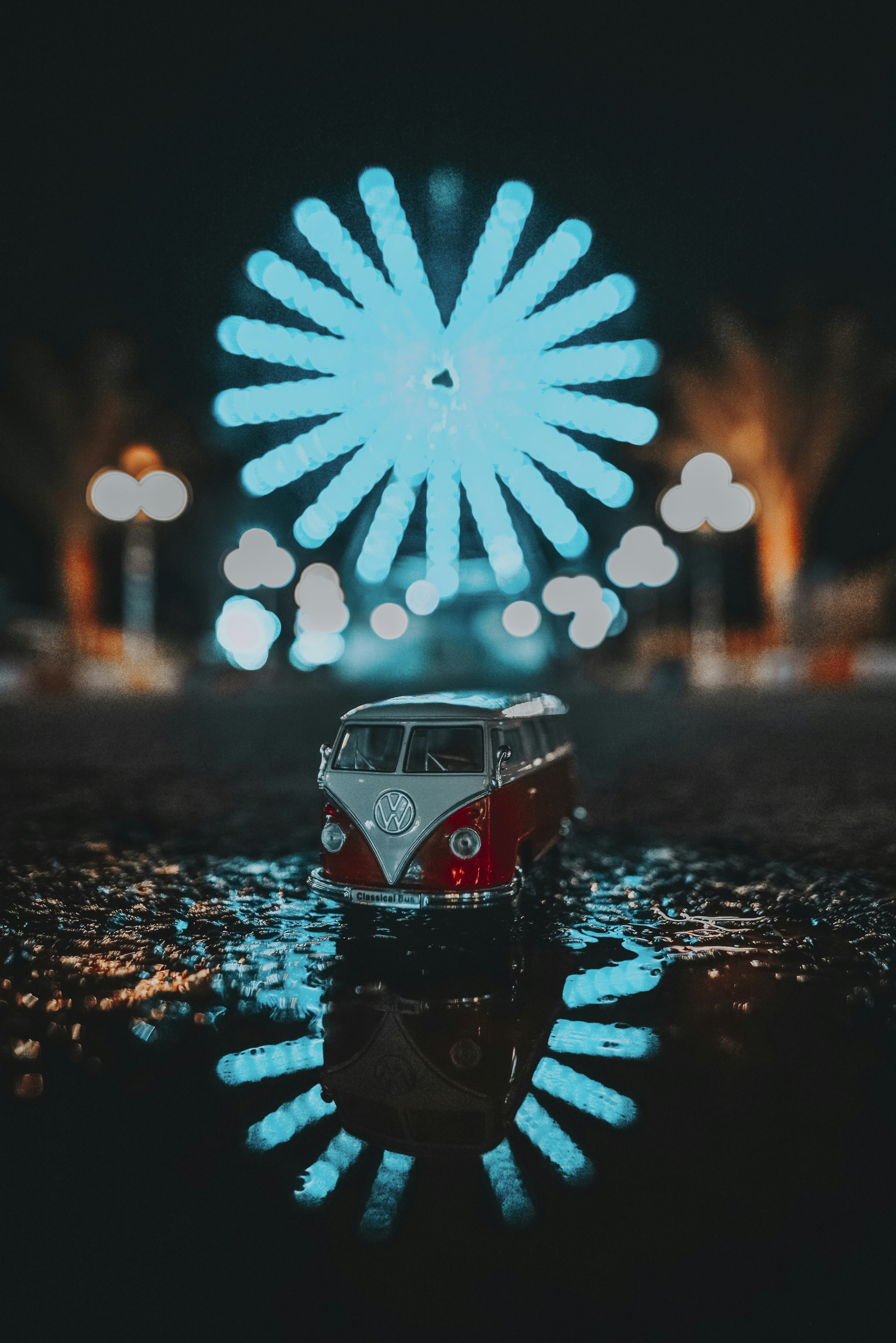 Miniature Volkswagen van reflecting in a puddle with a vibrant ferris wheel backdrop at night.