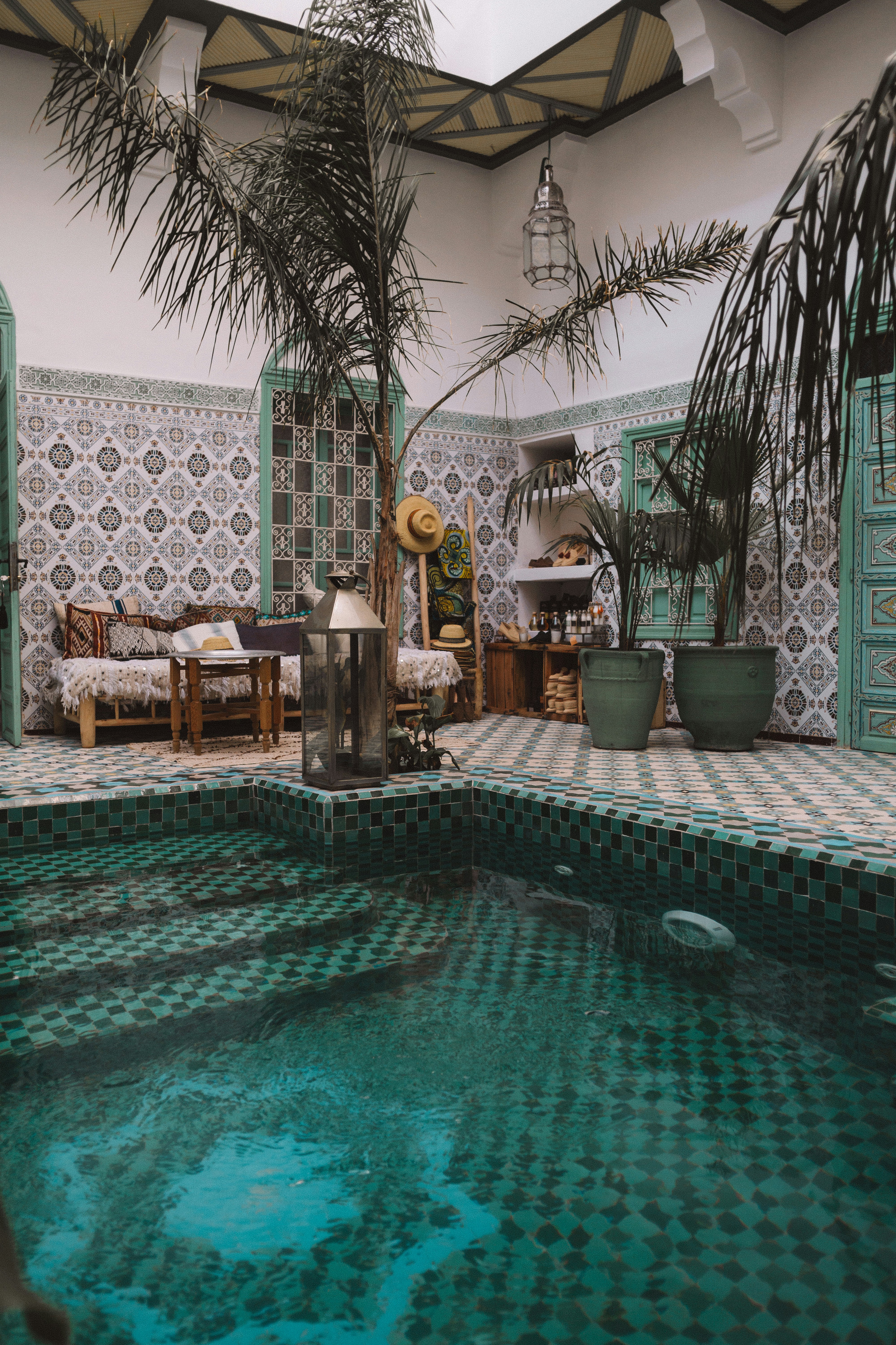 A tranquil indoor courtyard featuring a patterned tiled pool, lush greenery, and cozy seating, evoking a sense of relaxation and cultural charm.