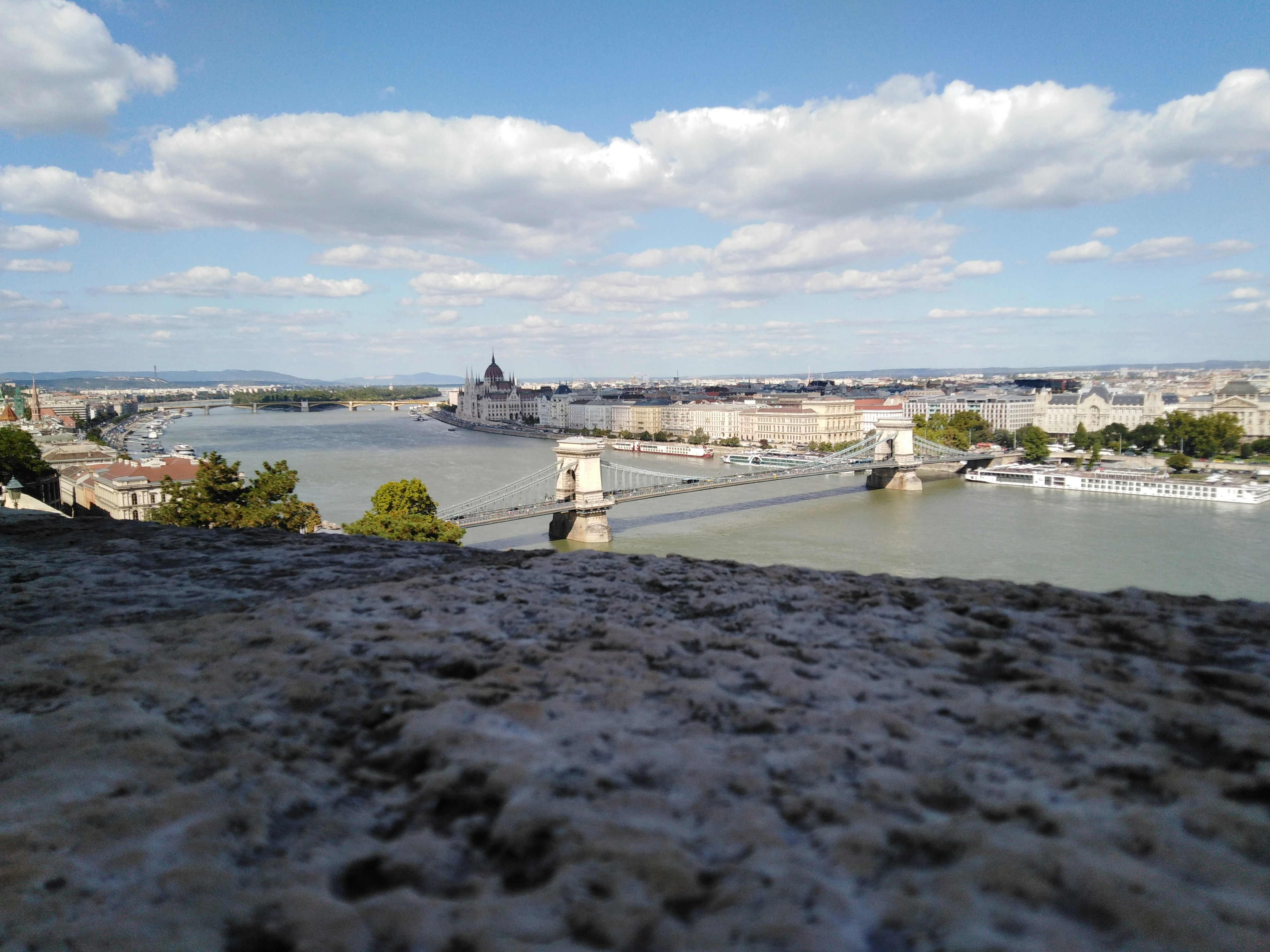 View of the Chain Bridge connecting Buda and Pest, with the Parliament building in the background under a cloudy sky.