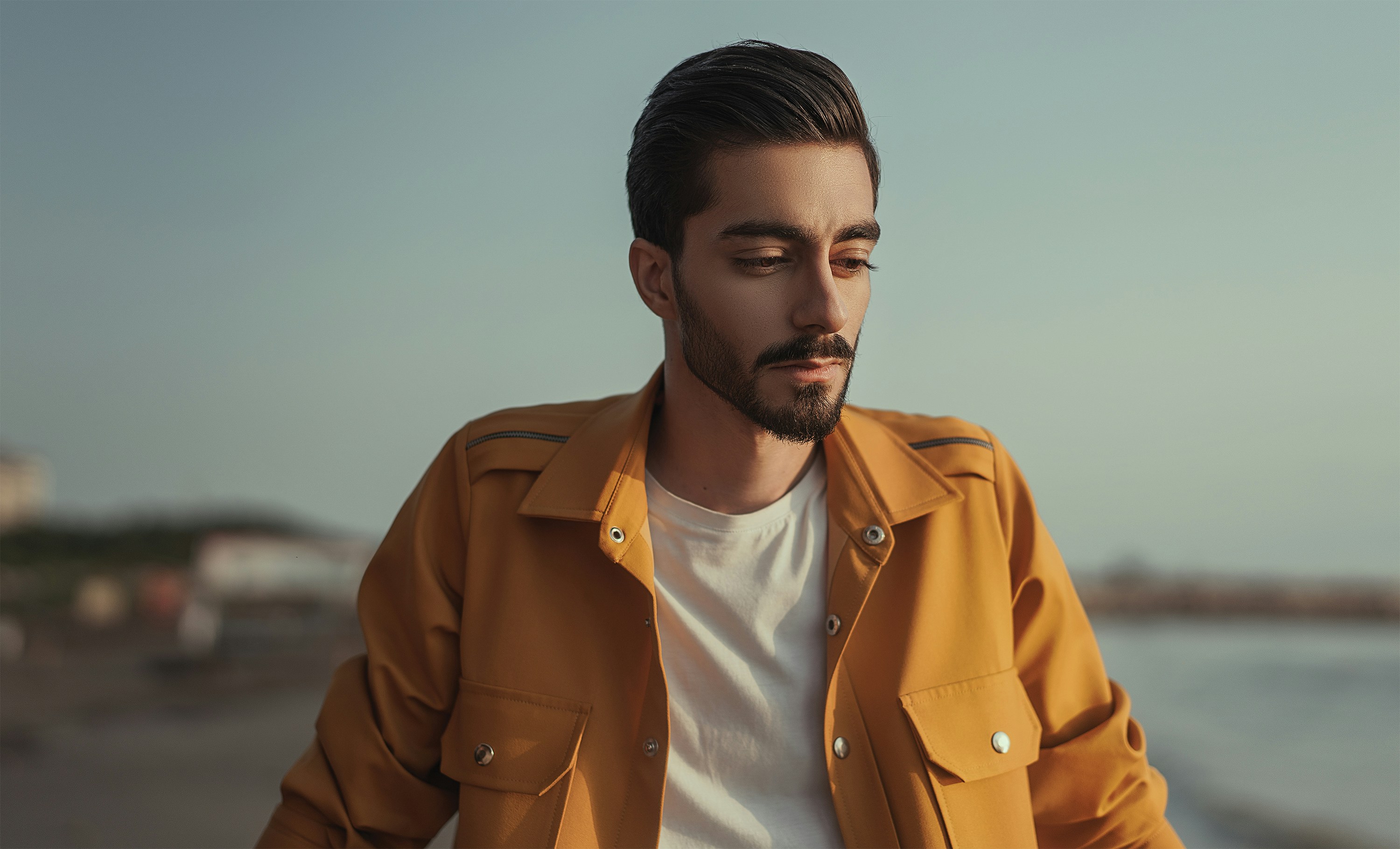 Man in a mustard jacket stands on a beach with a serene expression under a clear sky.