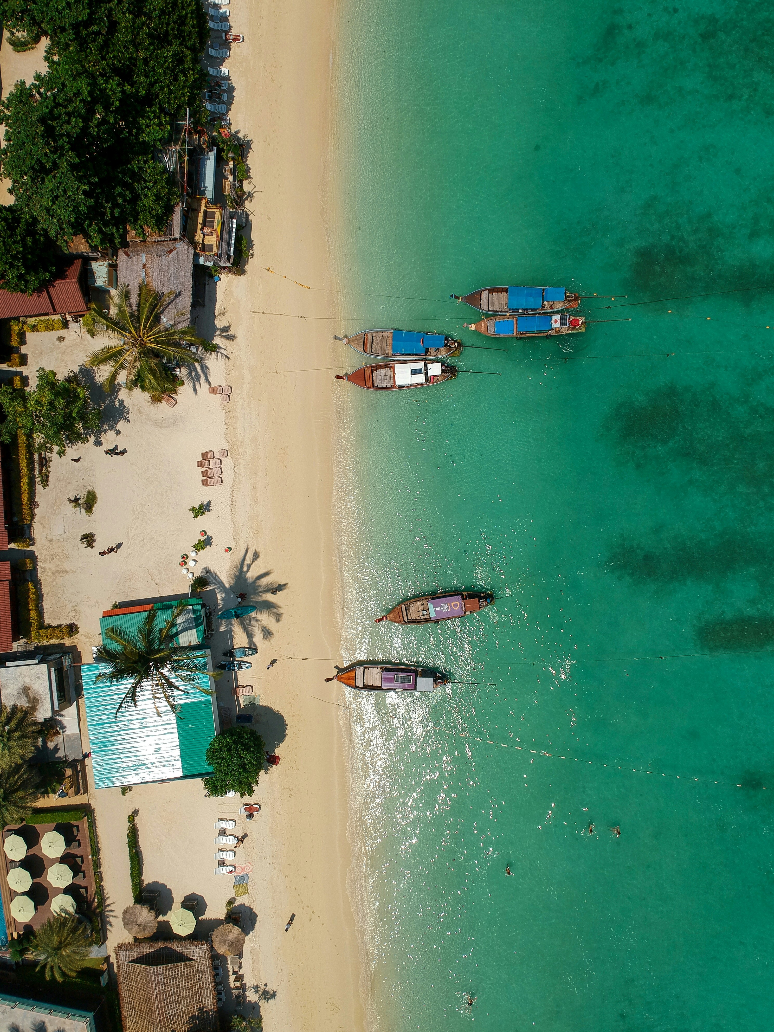 Aerial view of a serene beach with vibrant boats anchored in clear turquoise waters, framed by lush greenery and sandy shores.