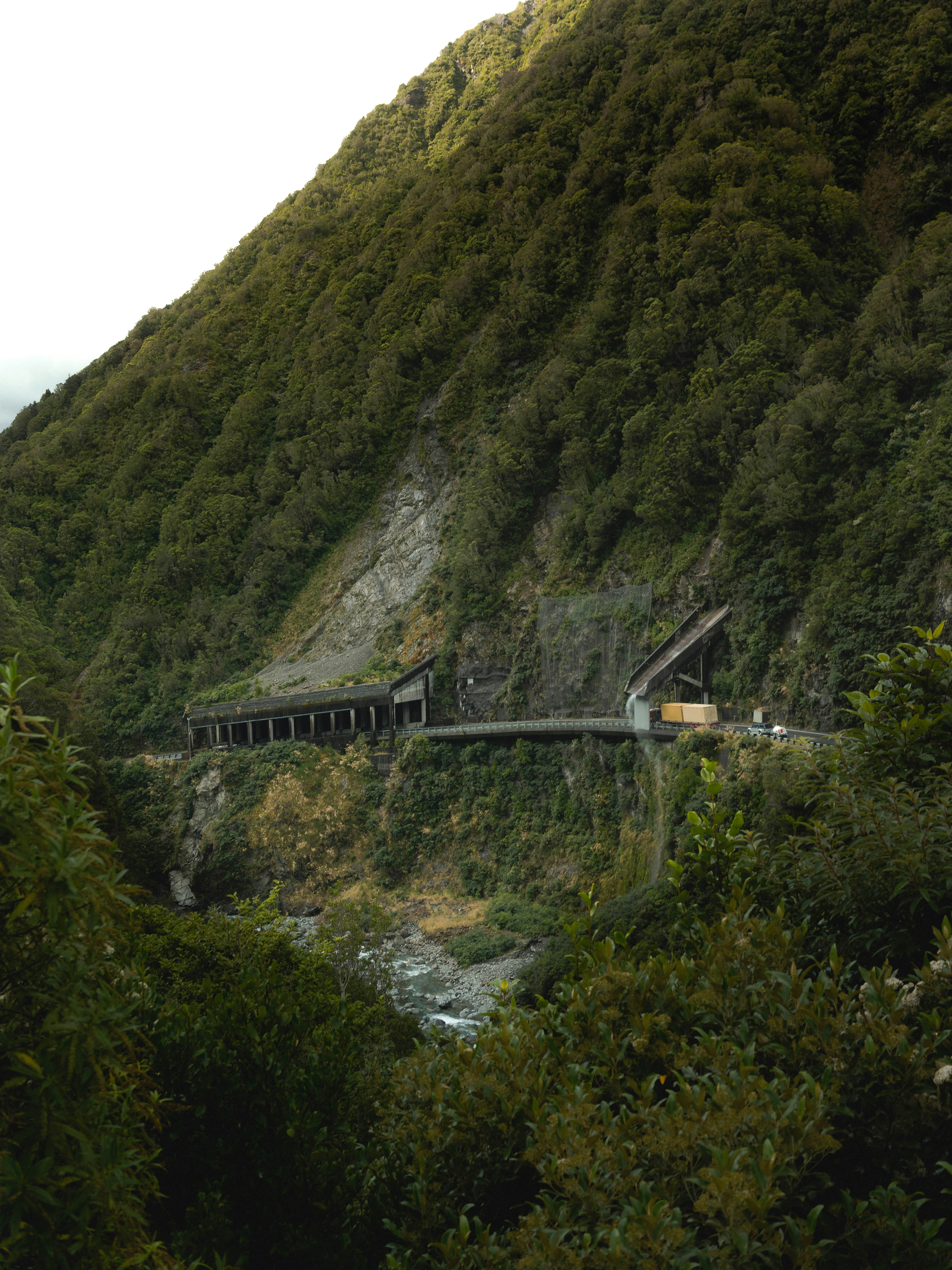 Abandoned industrial structure nestled against a steep mountain, surrounded by lush greenery and a winding river below.