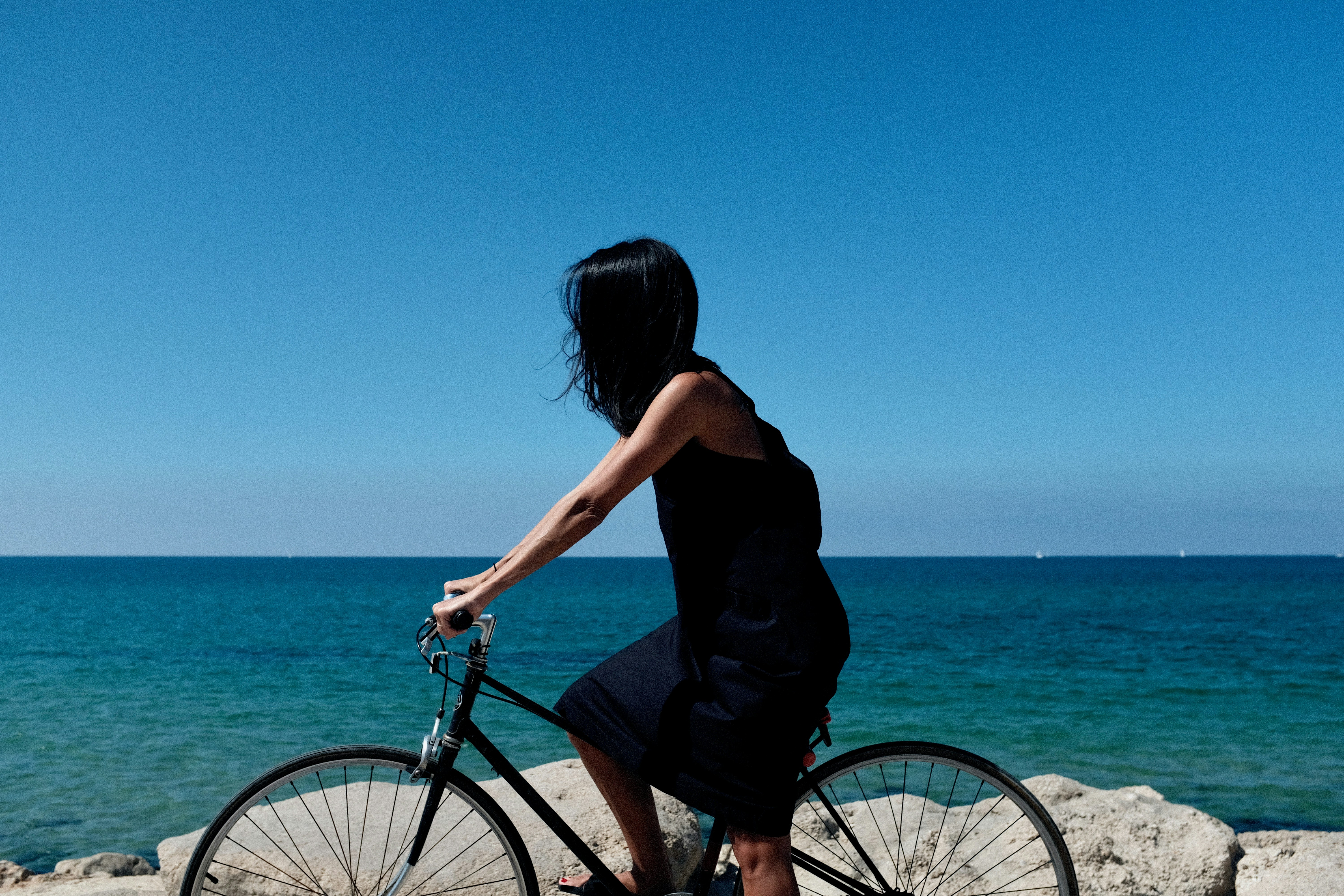Woman on a bicycle by the sea under a clear blue sky.