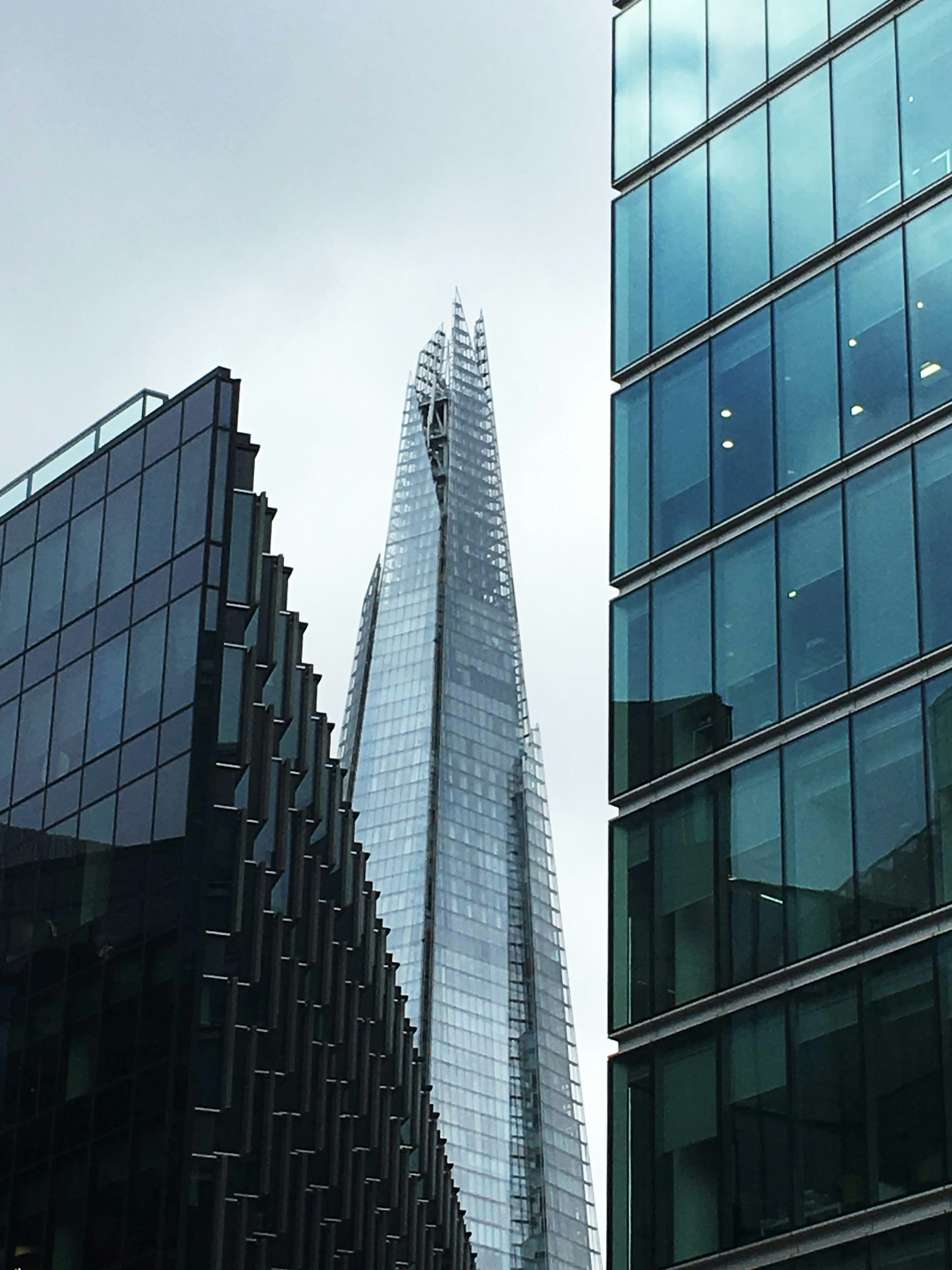 The Shard rises majestically between contemporary glass buildings, showcasing its unique design against a cloudy sky.