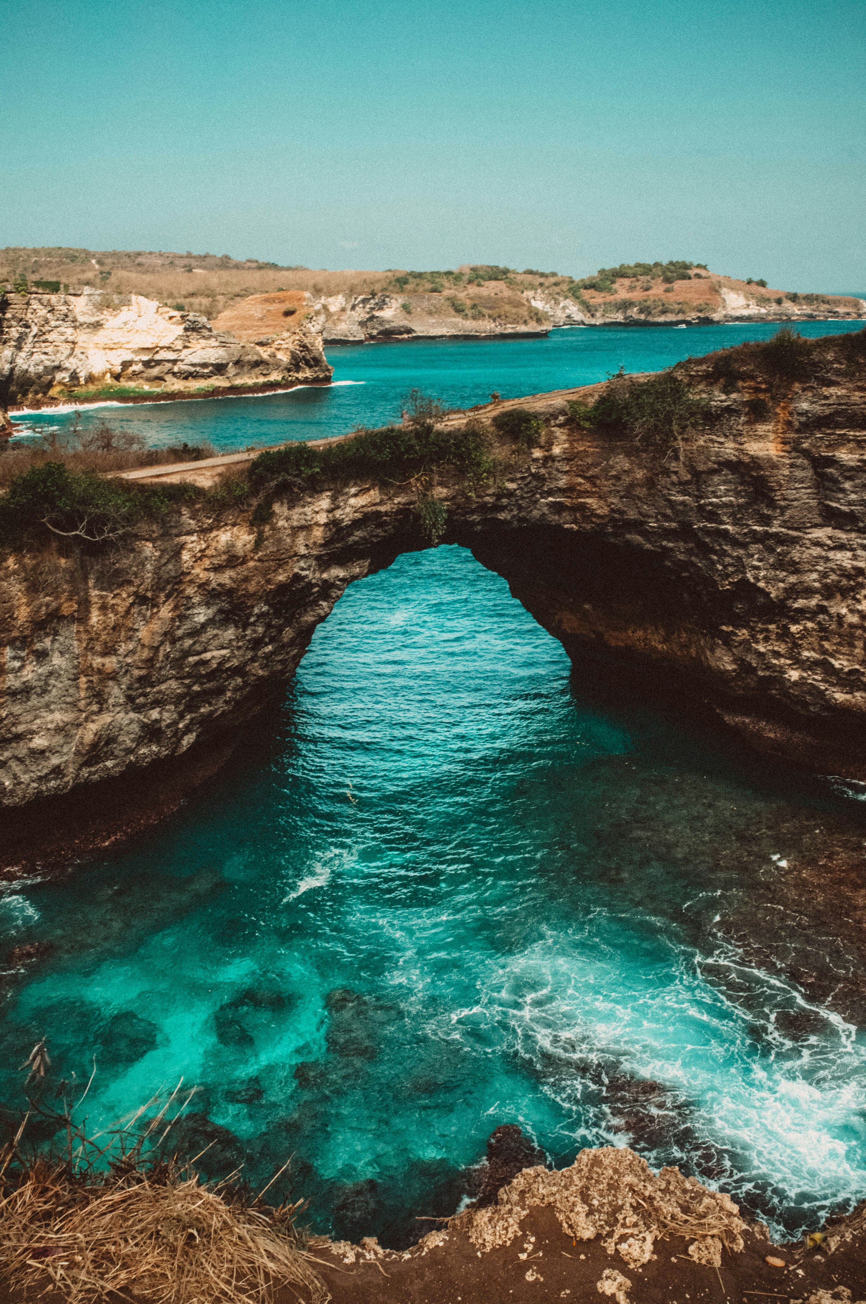 Rock formation arching over vibrant turquoise sea with distant cliffs under a clear sky.